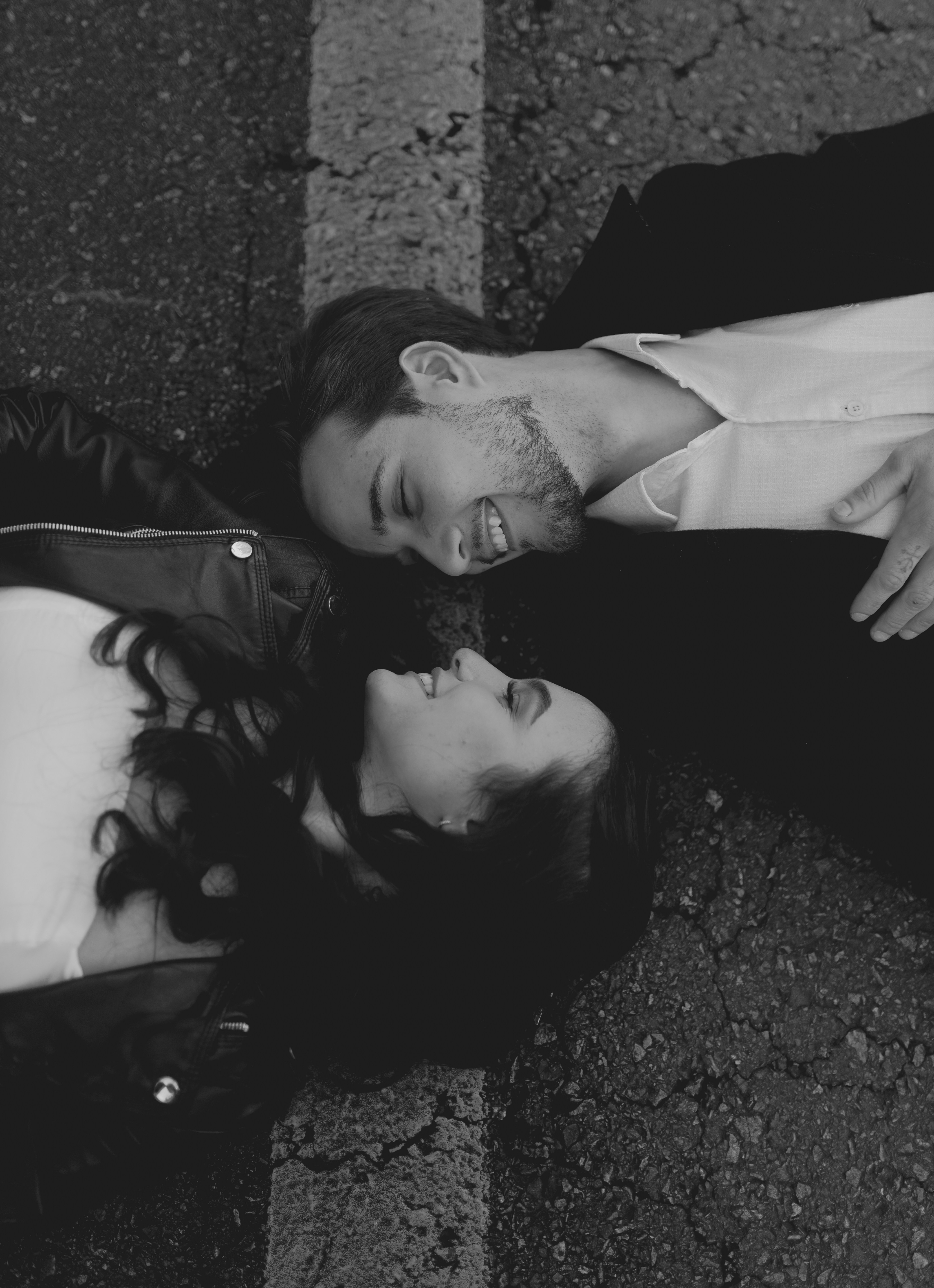 Couple lying on a cracked road, sharing a moment of laughter and connection. The contrasting textures of their clothing and the ground enhance the scene's intimacy.