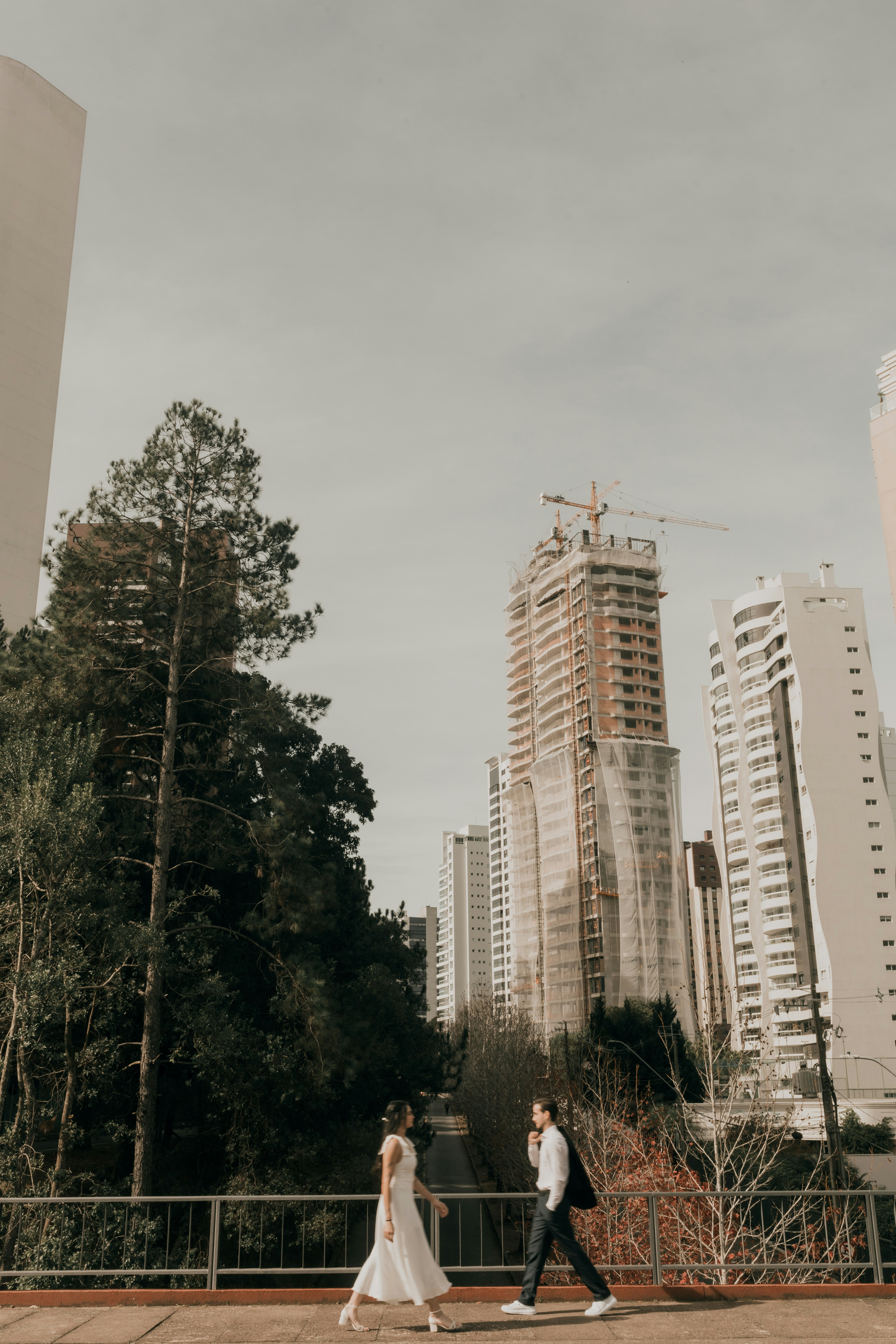 Couple strolling through an urban landscape, framed by towering buildings and lush greenery. The scene captures the blend of nature and modern architecture.