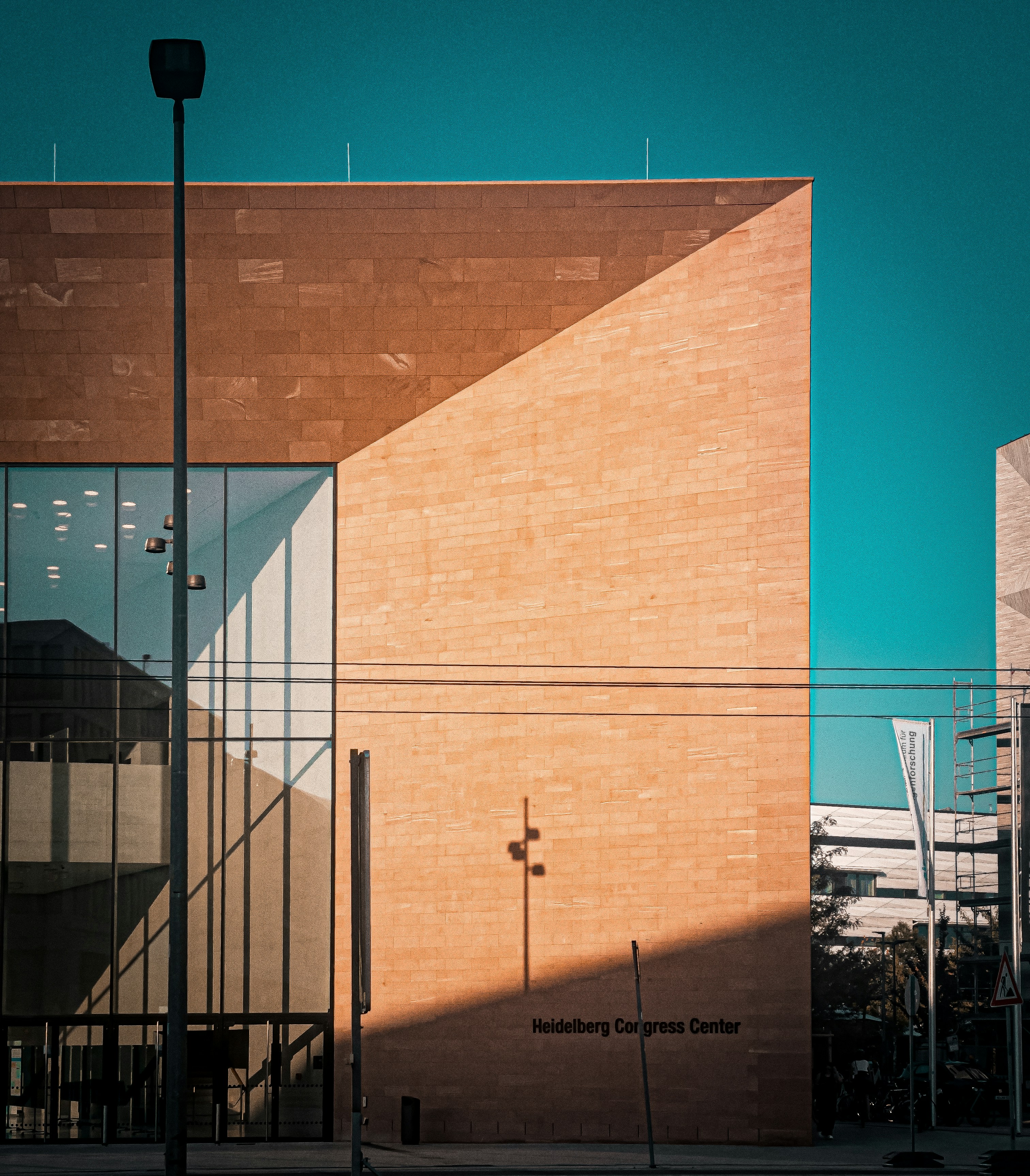 A beige stone and brick facade of the Heidelberg Congress Center featuring a large glass entrance on the left. A tall lamppost casts a shadow across the building’s surface, with overhead utility wires. Text reading “Heidelberg Congress Center” is visible on the right side of the facade, and a public walkway runs along the front. | A modern building with a diagonal shadow.