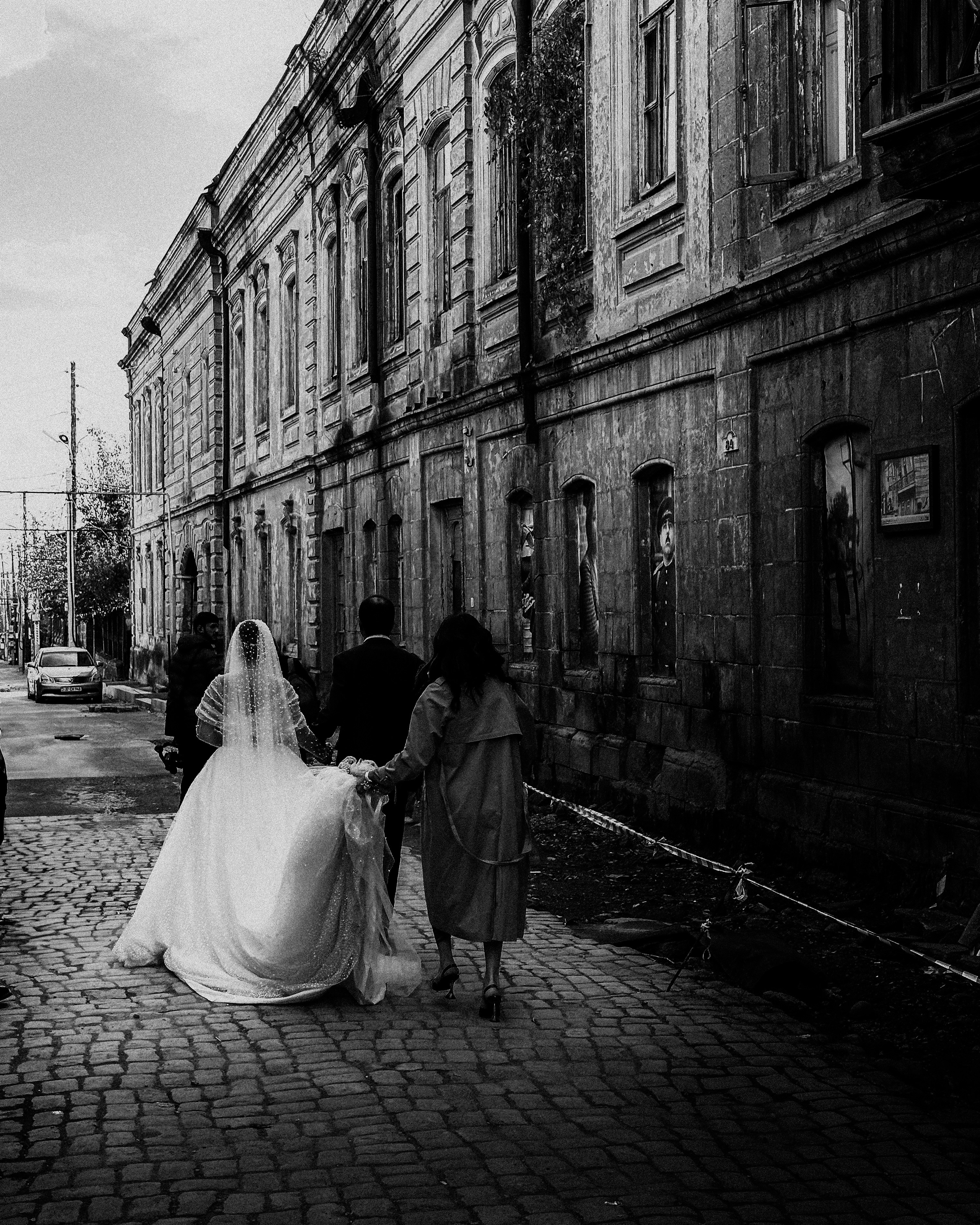 Bride in a flowing gown walks along a cobblestone street, accompanied by two figures, against a backdrop of historic architecture.