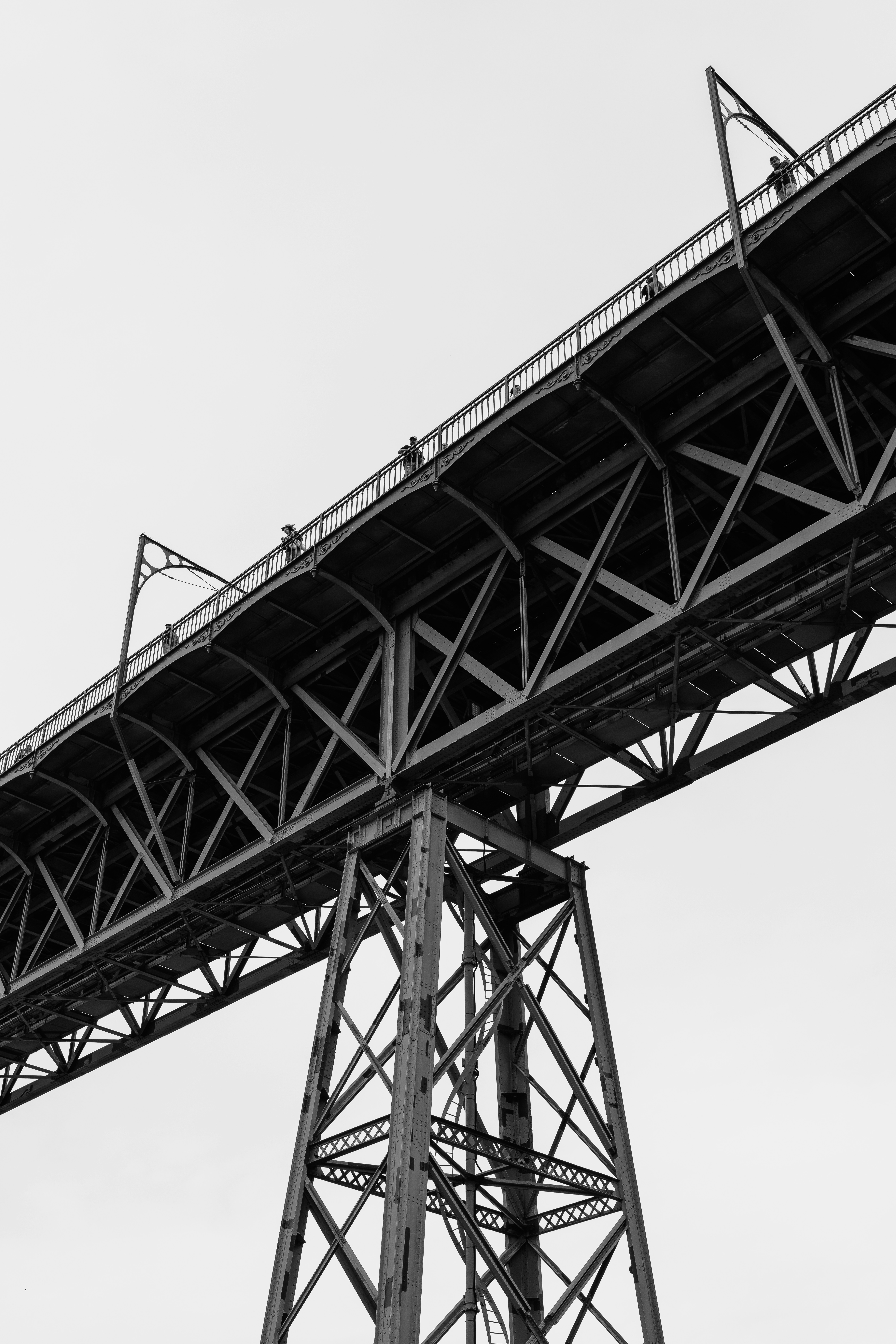 A monochrome view of a towering bridge from below, showcasing its intricate steel framework and the pedestrians above.