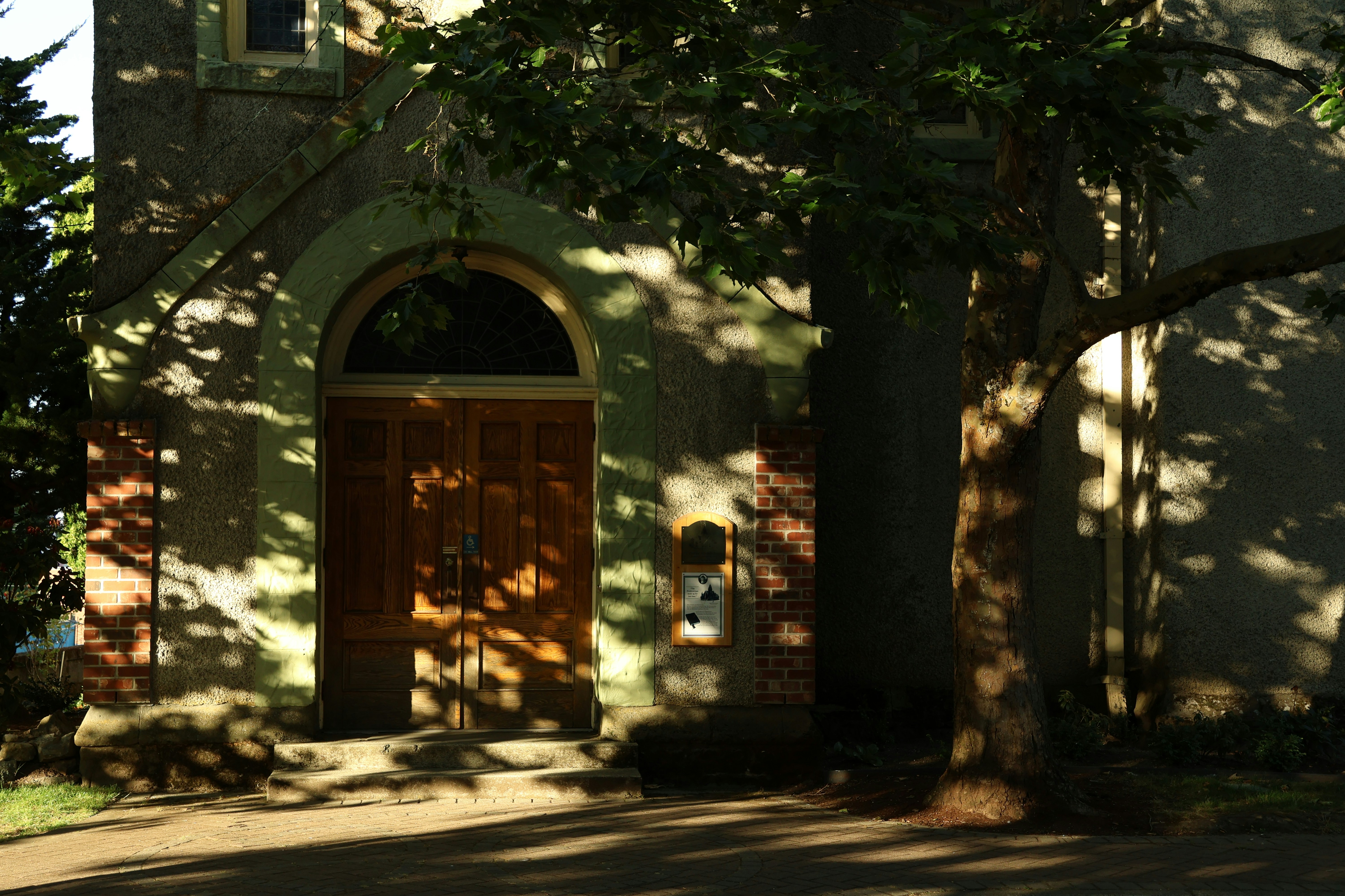 Historic wooden doors framed by intricate shadows cast by surrounding trees, inviting exploration. A subtle blend of nature and architecture.