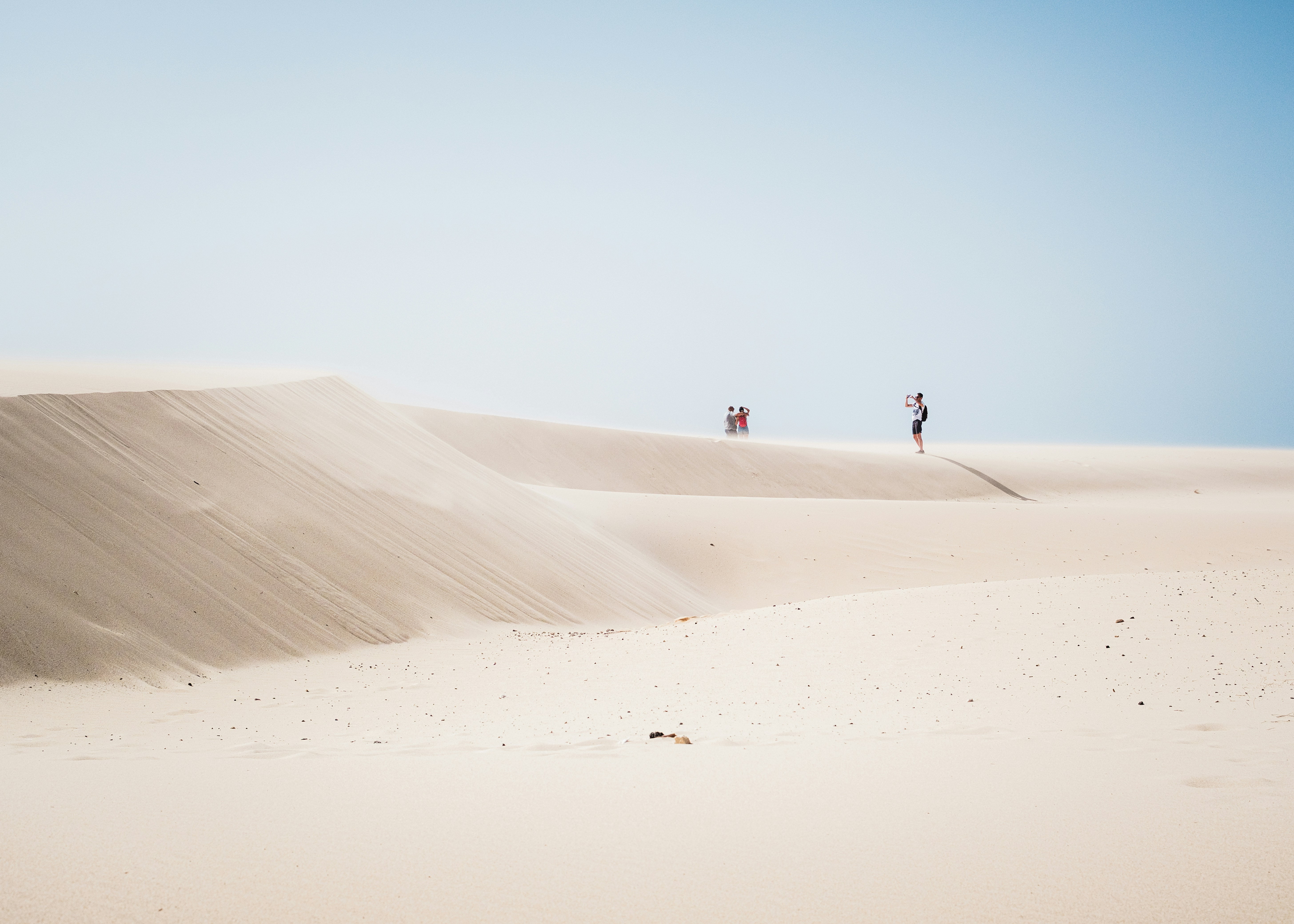 The image shows the Bolonia Dune north of Tarifa. Bright, fine sand forms large, gently rolling hills. Three people stand on the crest of the dune under a clear blue sky, one of them photographing the landscape. There is no visible vegetation; everything appears vast and calm. | People traverse the sand dunes under a bright sky.