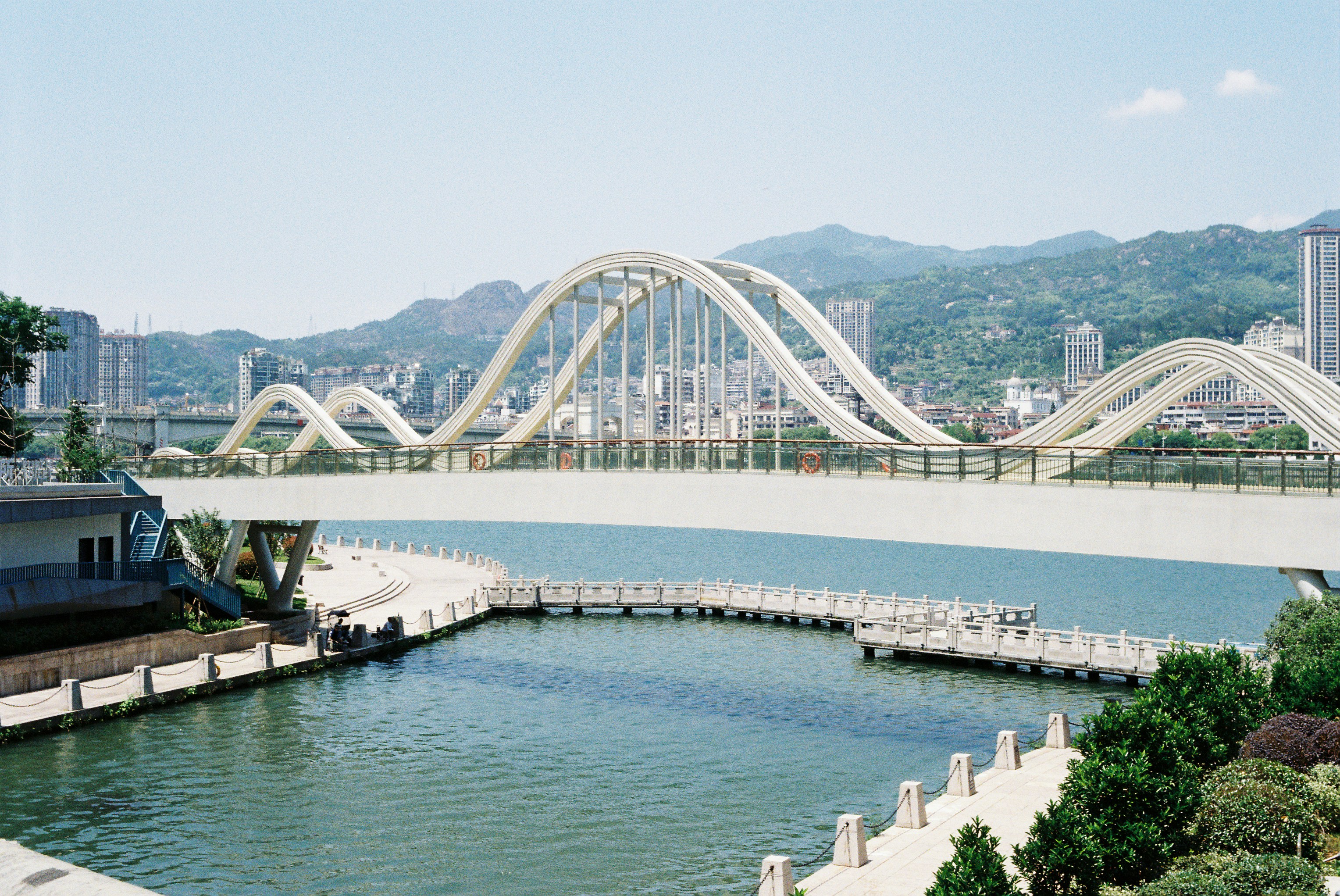 A modern bridge arches gracefully over a serene body of water, framed by lush greenery and urban structures in the background.