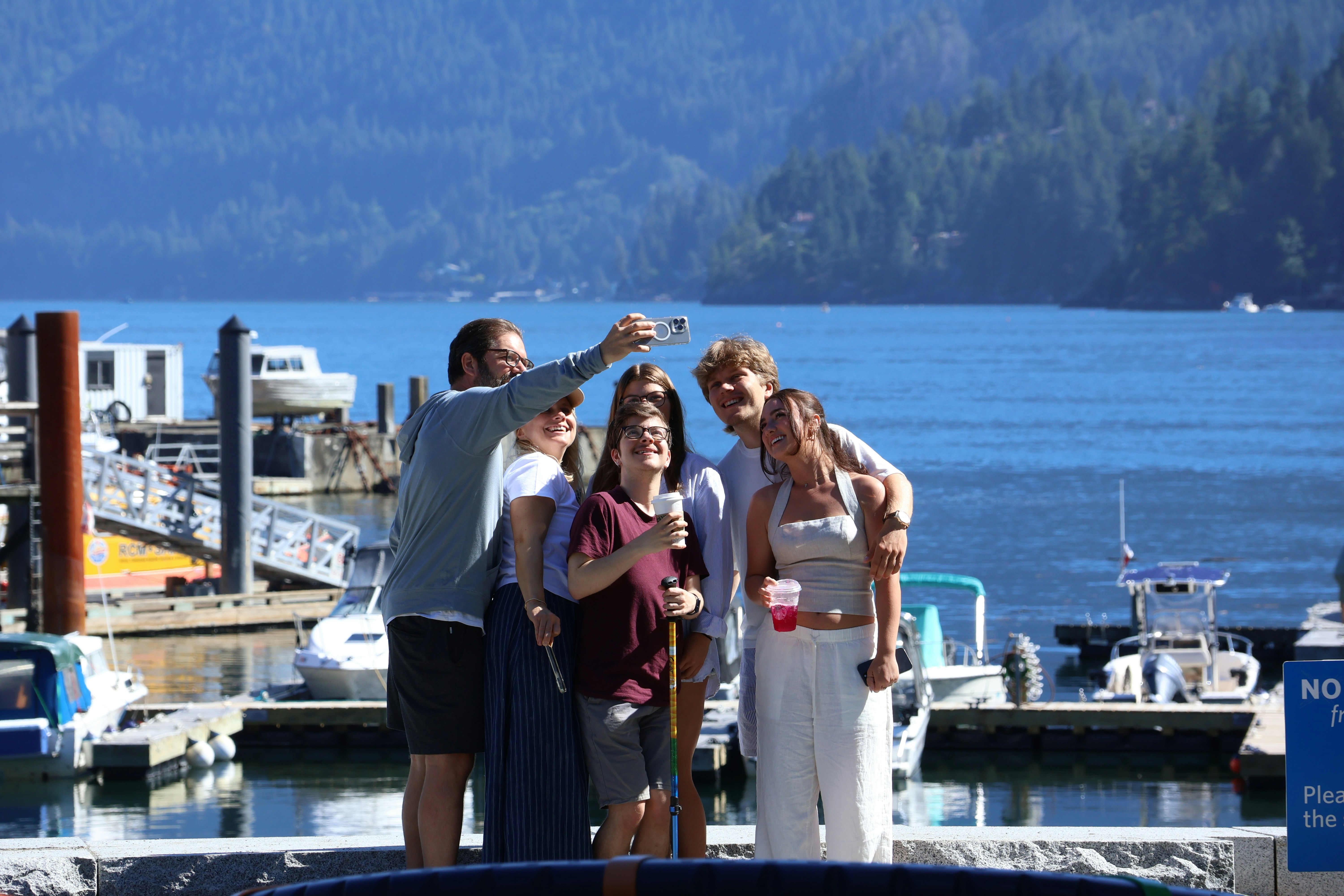 Group of friends taking a selfie on a sunny day near a marina, with boats and mountains in the background.