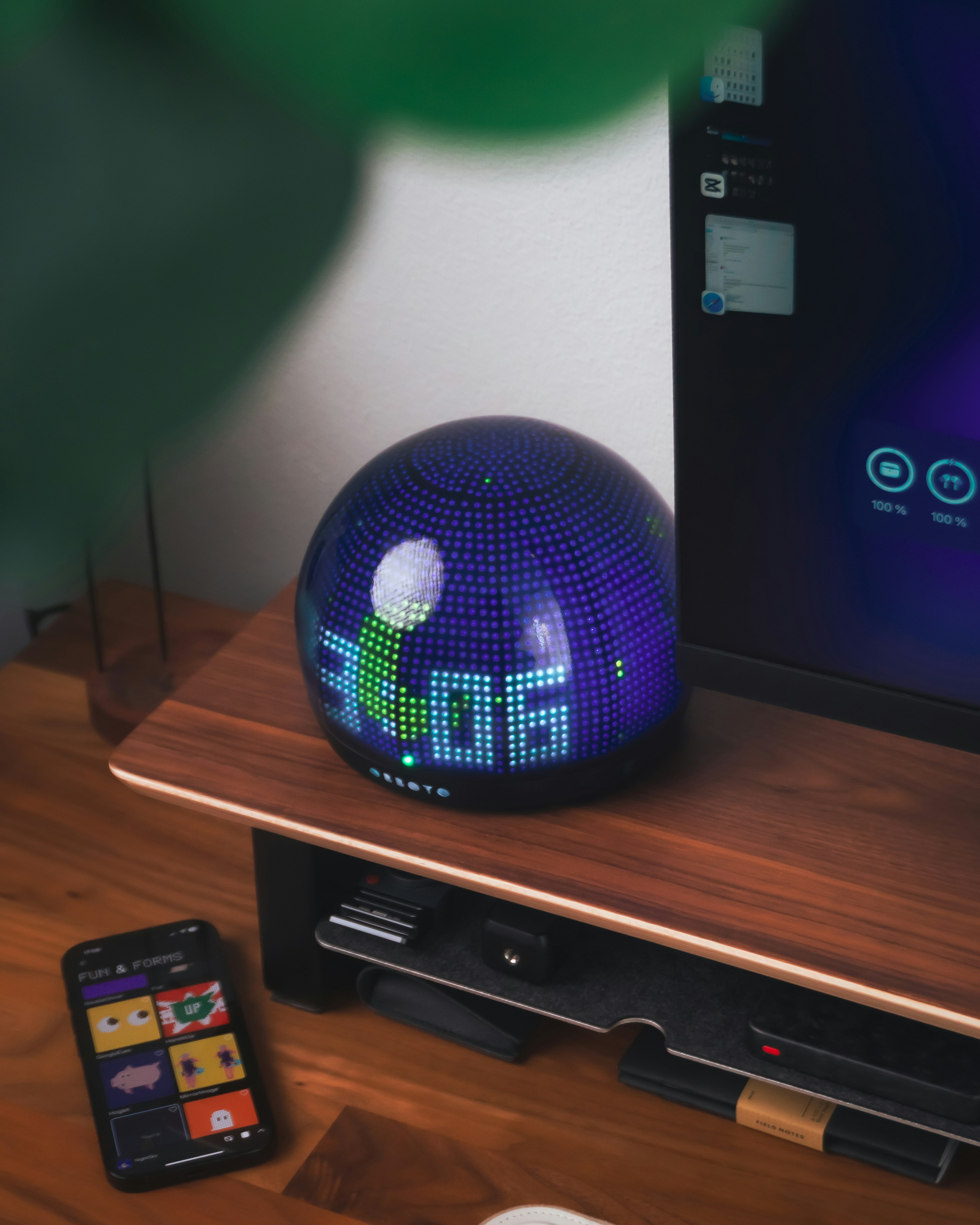 A vibrant, illuminated speaker displaying digital time on a wooden desk, accompanied by a smartphone and a glimpse of a monitor in the background.
