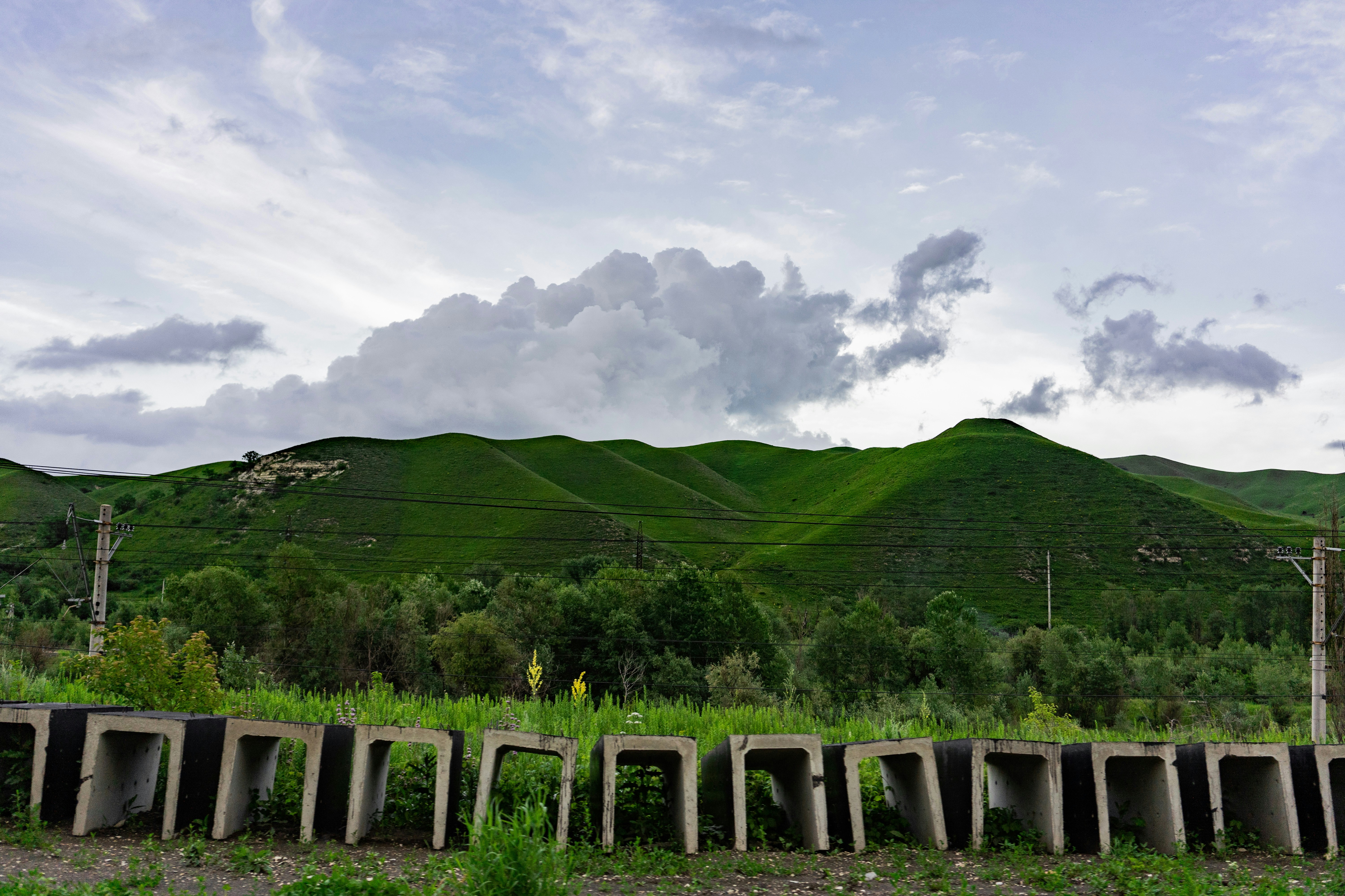 Vibrant green hills undulate beneath a dramatic sky, framed by concrete structures in the foreground. The scene captures the harmony between man-made elements and natural beauty.