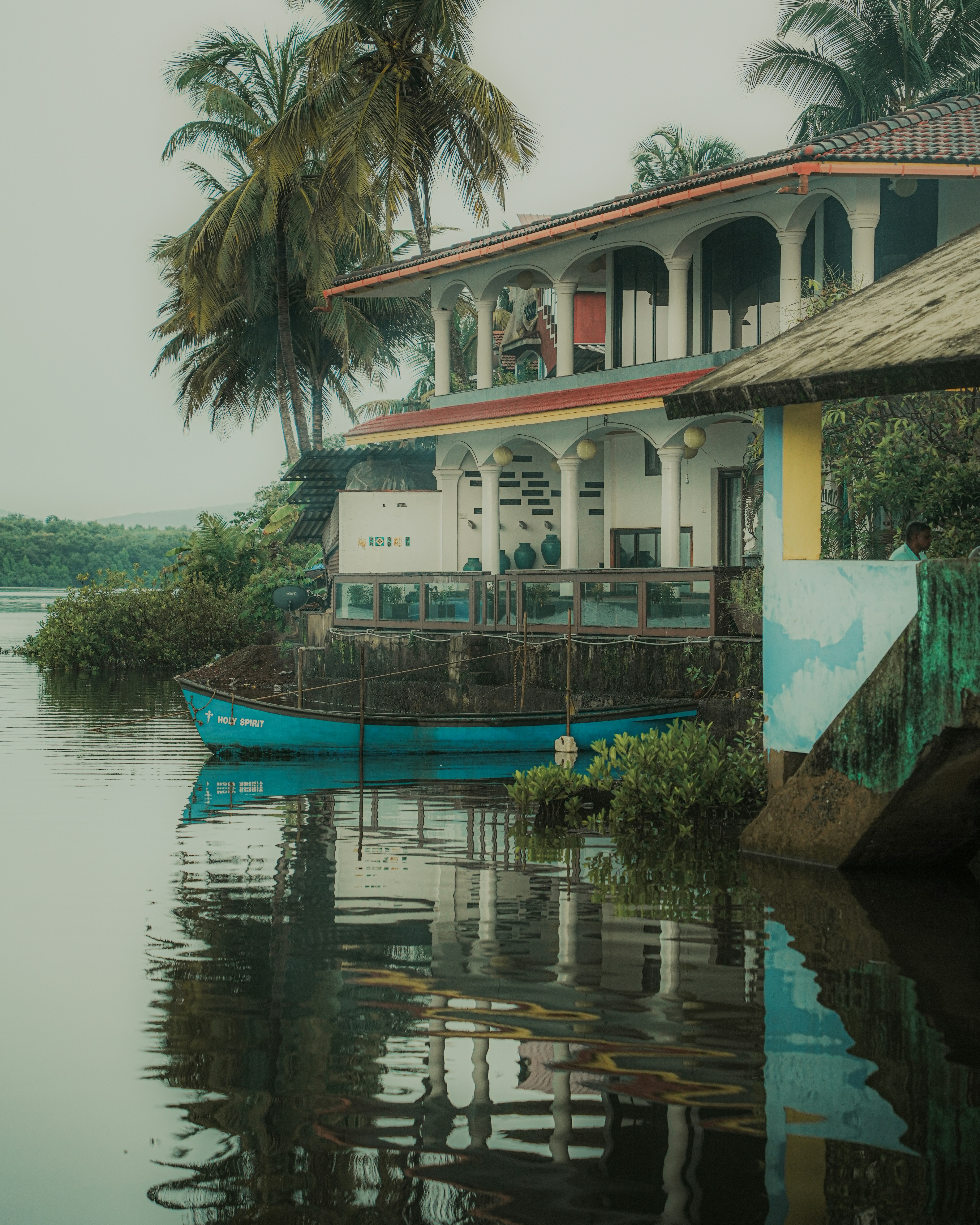 Charming waterside house with a blue boat anchored nearby, surrounded by lush greenery and palm trees, reflecting in the calm water.