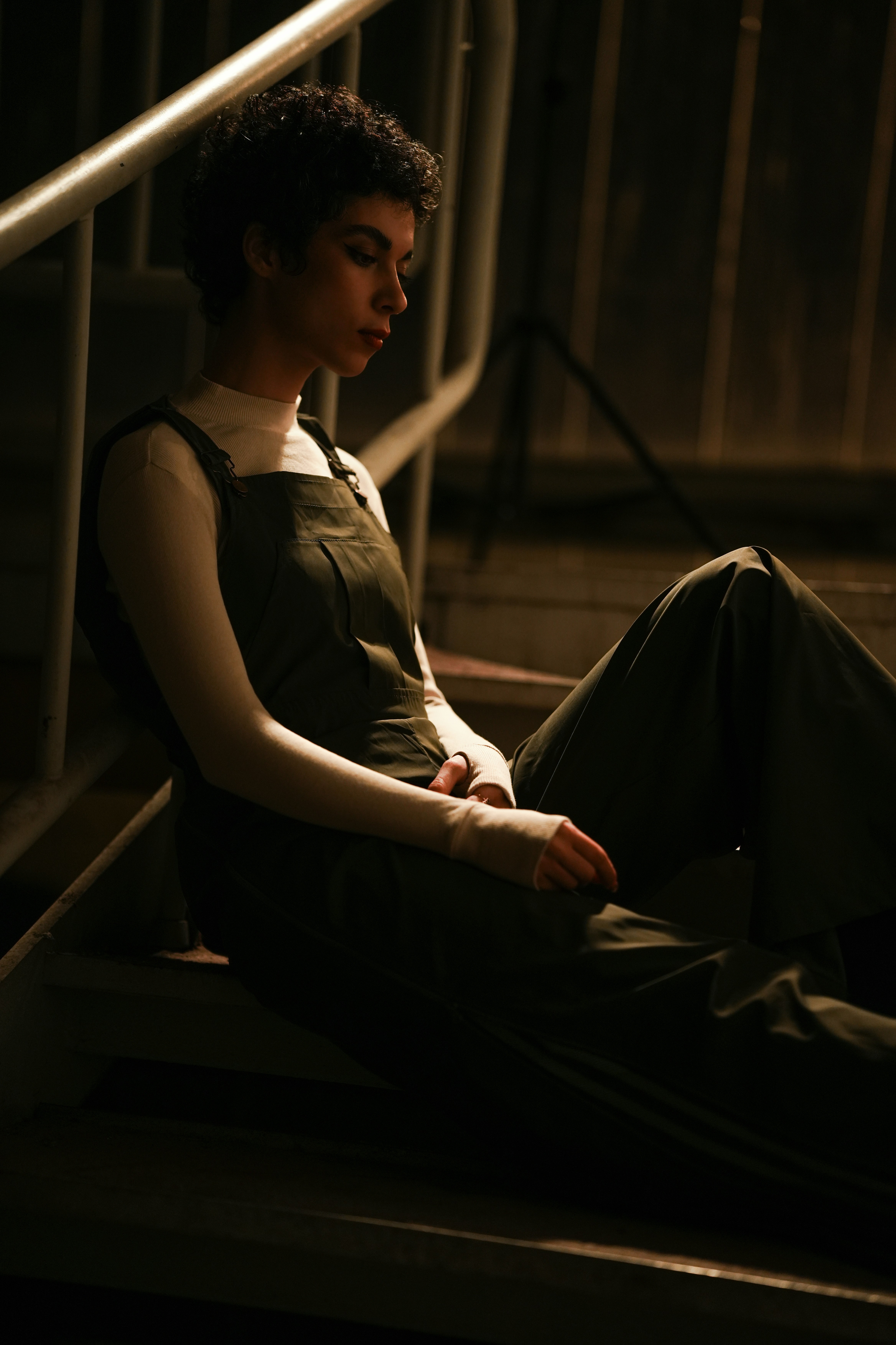 A contemplative figure seated on stairs, dressed in a dark outfit, immersed in soft light and shadows.