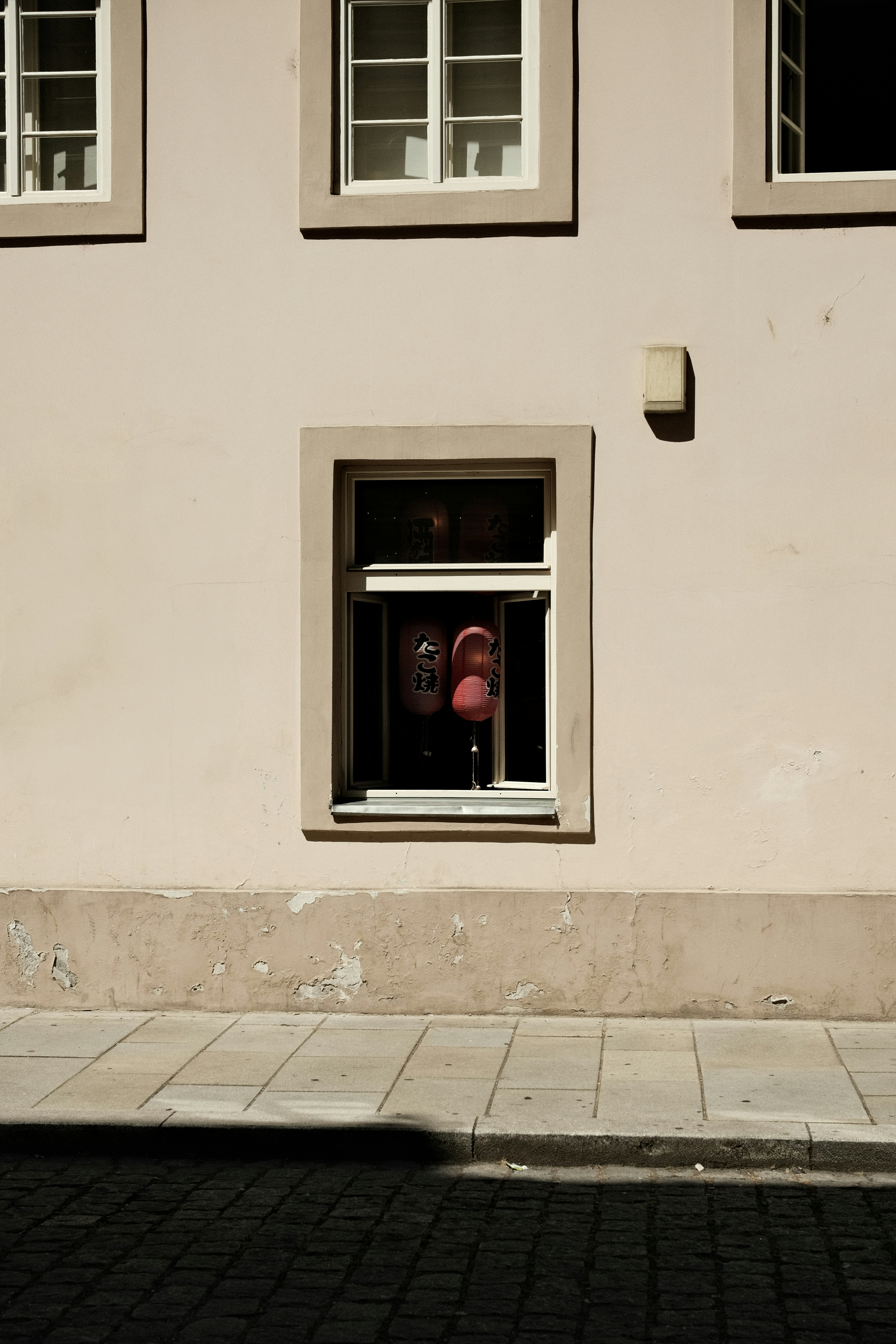 A window is framed by a light-colored wall. Inside, two red paper lanterns are visible, adding contrast to the neutral exterior. | A building's windows with a quirky display.