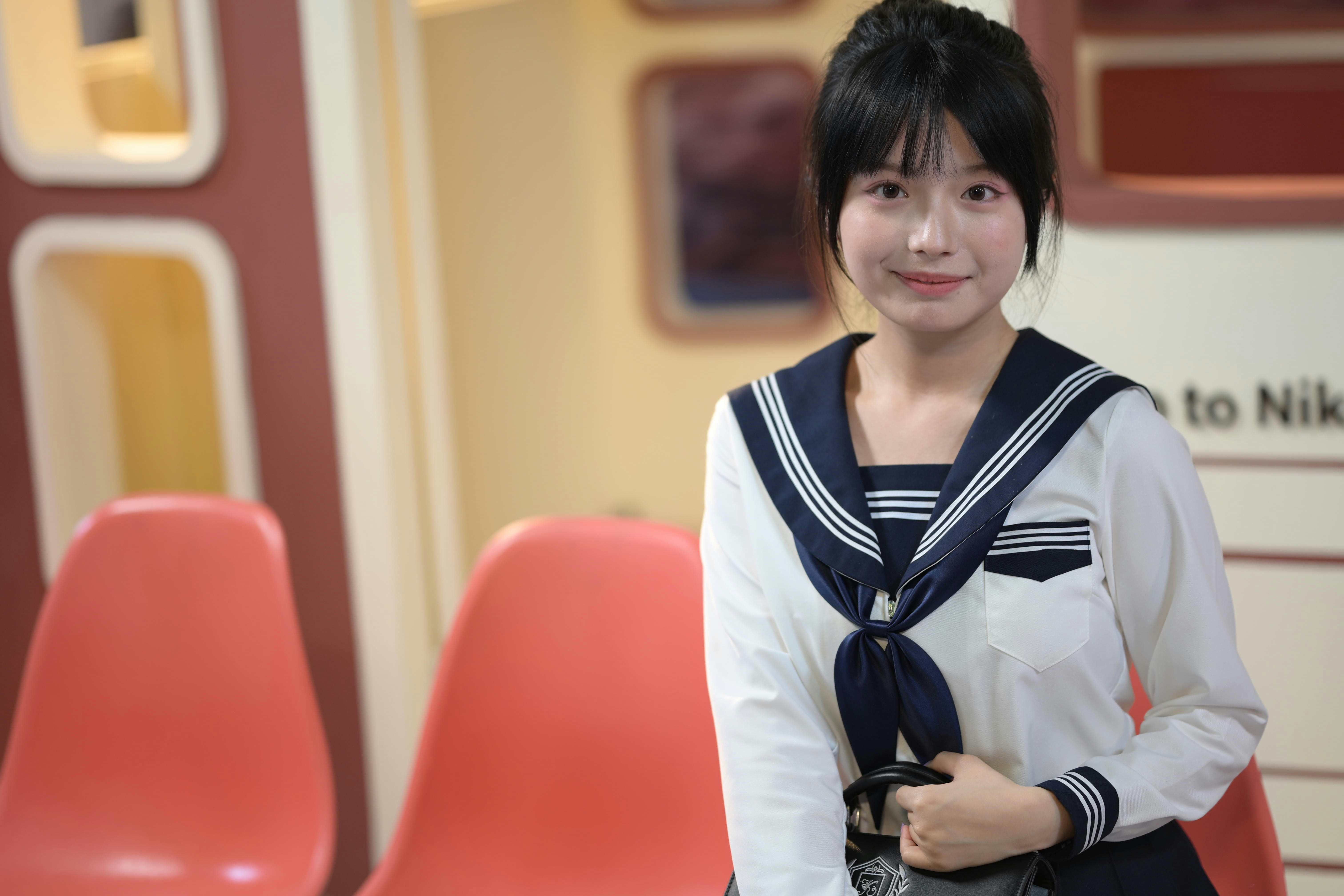 Young woman in a classic sailor uniform poses in a contemporary setting with vibrant red chairs.