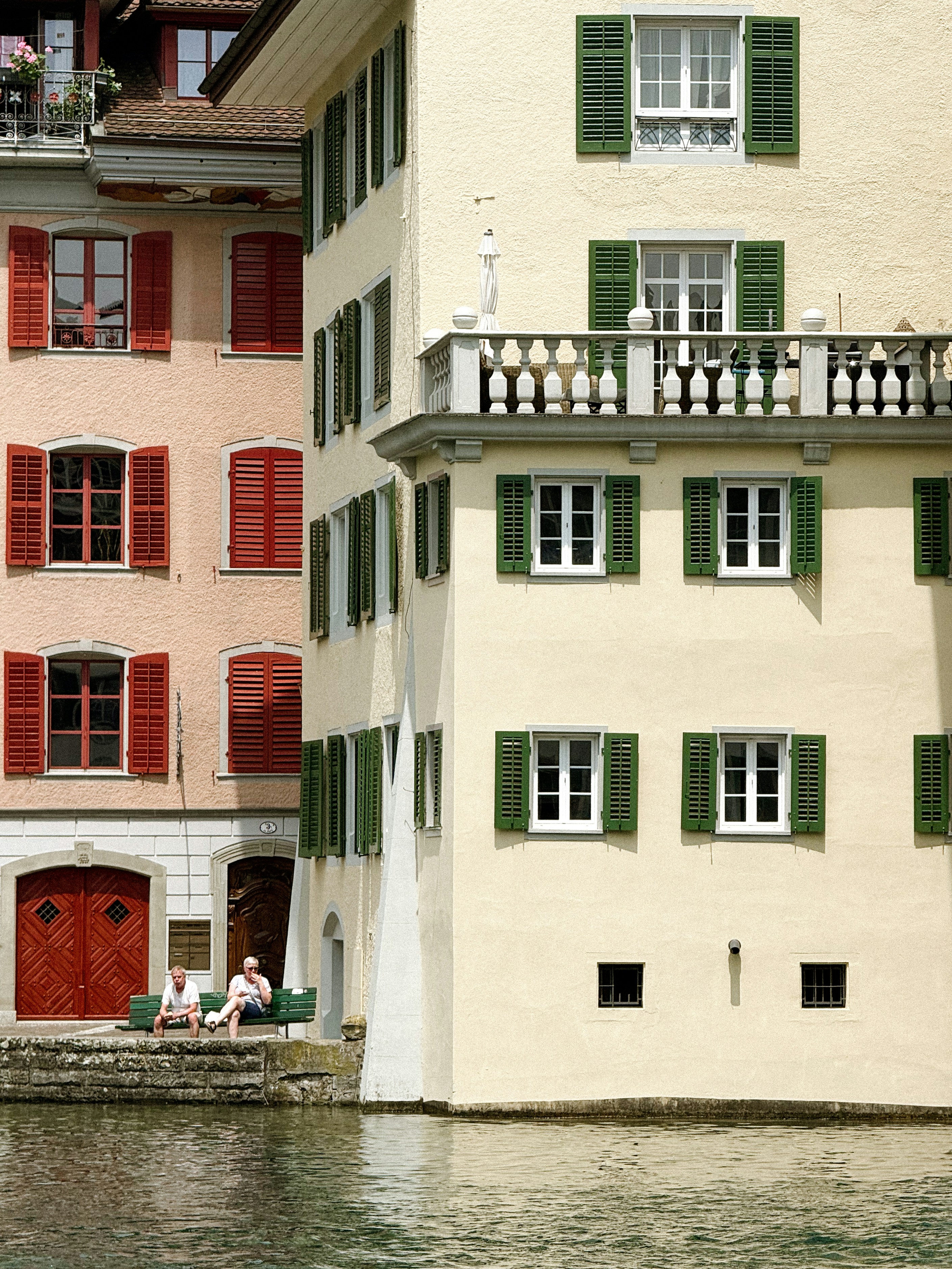 Two individuals relaxing on a bench by the lake, framed by pastel-colored buildings with vibrant green shutters. The serene waters reflect the charming architecture.