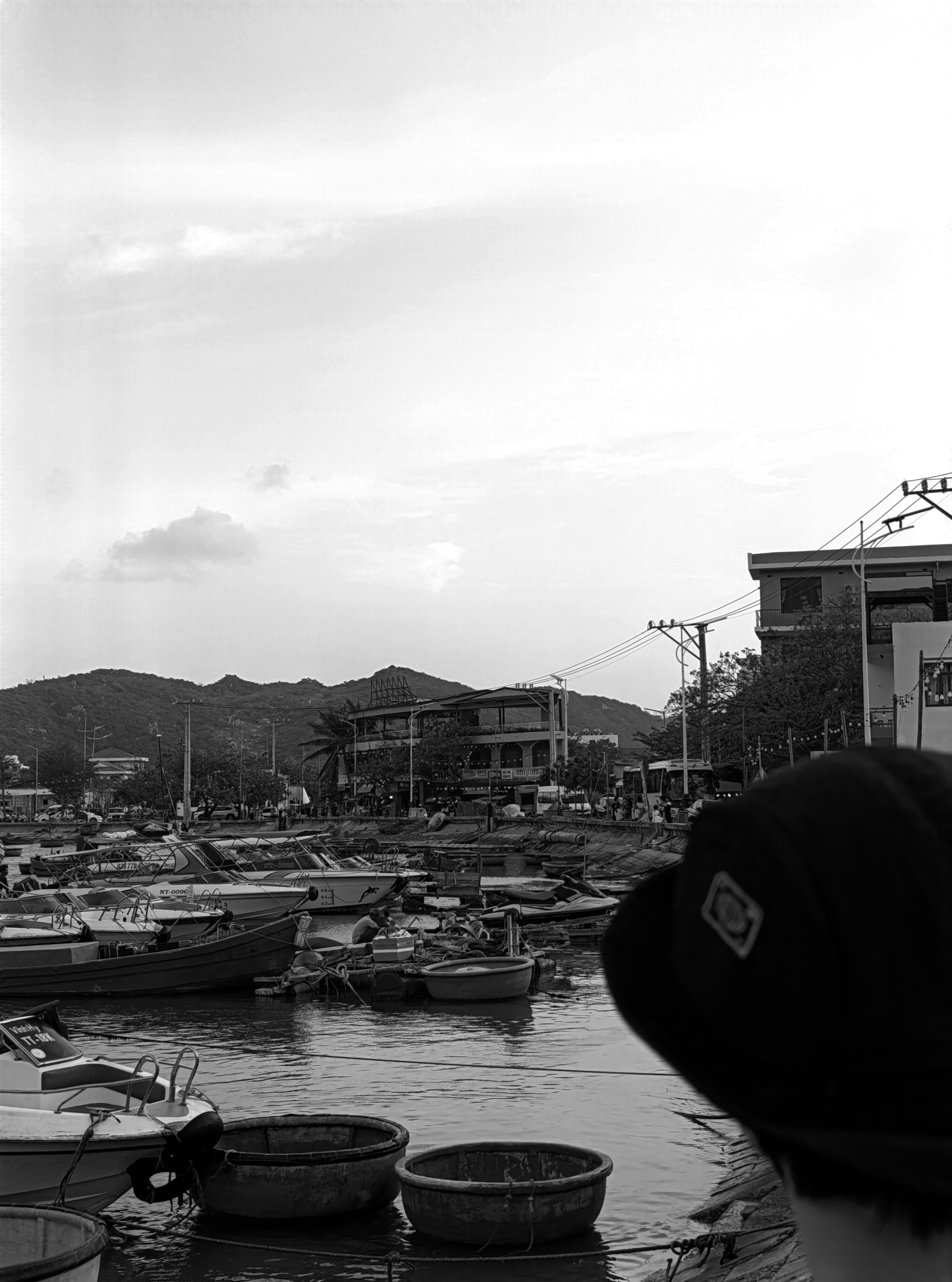 A tranquil harbor scene featuring boats and buildings, with a person partially visible in the foreground. The monochrome palette emphasizes the calmness of the setting.