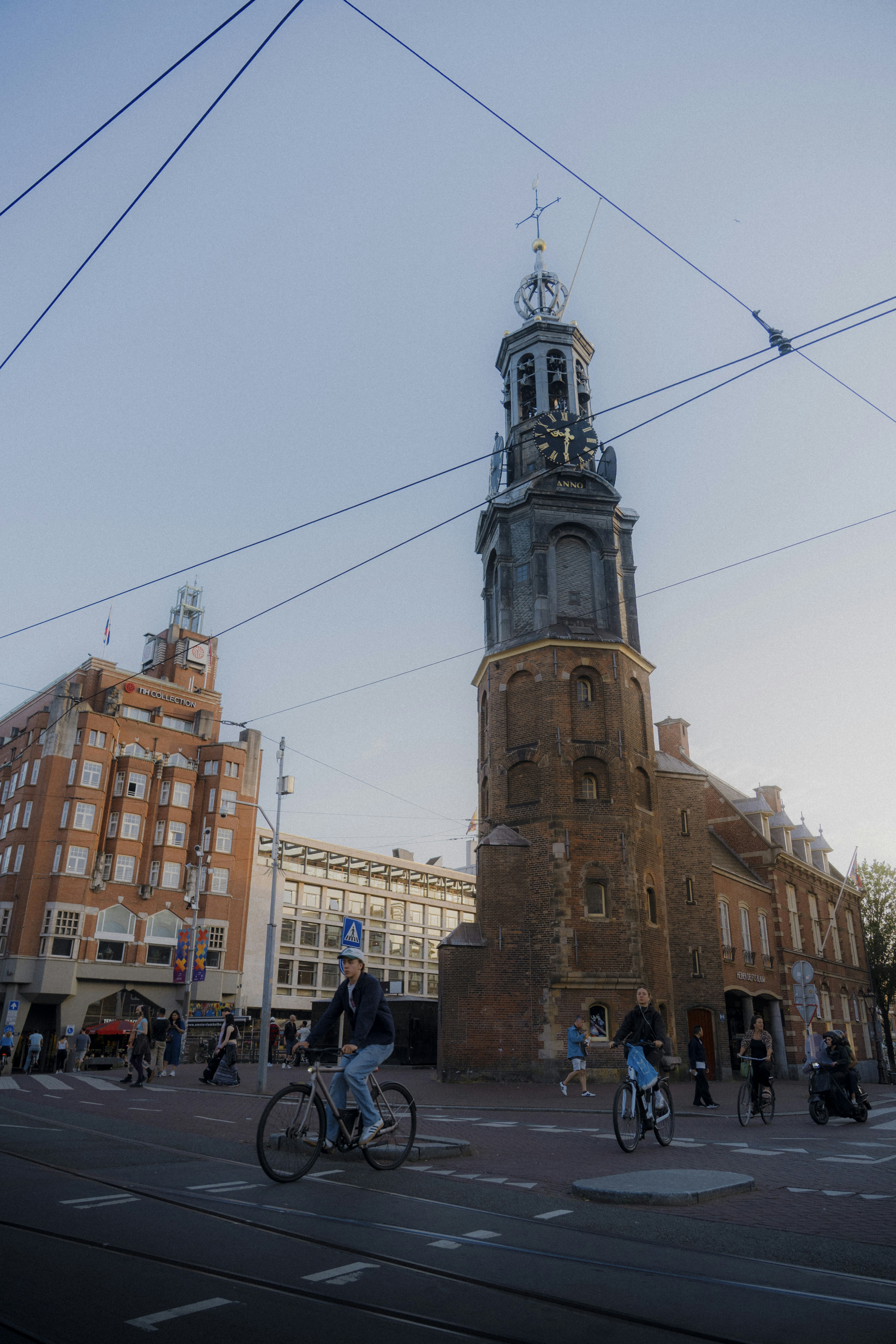 Historic clock tower stands prominently in a bustling square, surrounded by cyclists and pedestrians. The scene captures the essence of urban life.