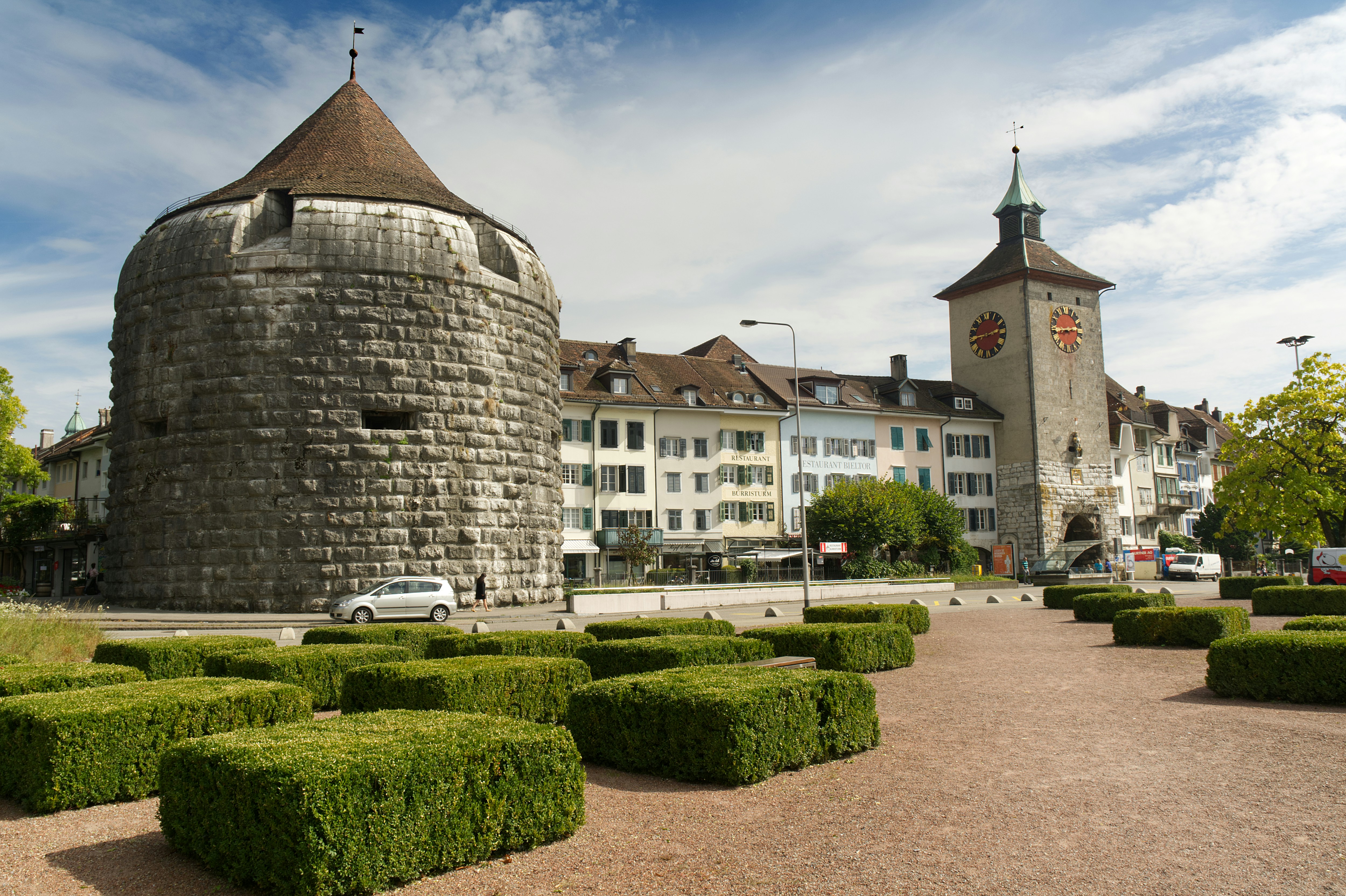 Historic stone tower and clock tower stand amidst manicured hedges and modern architecture, showcasing a blend of past and present.