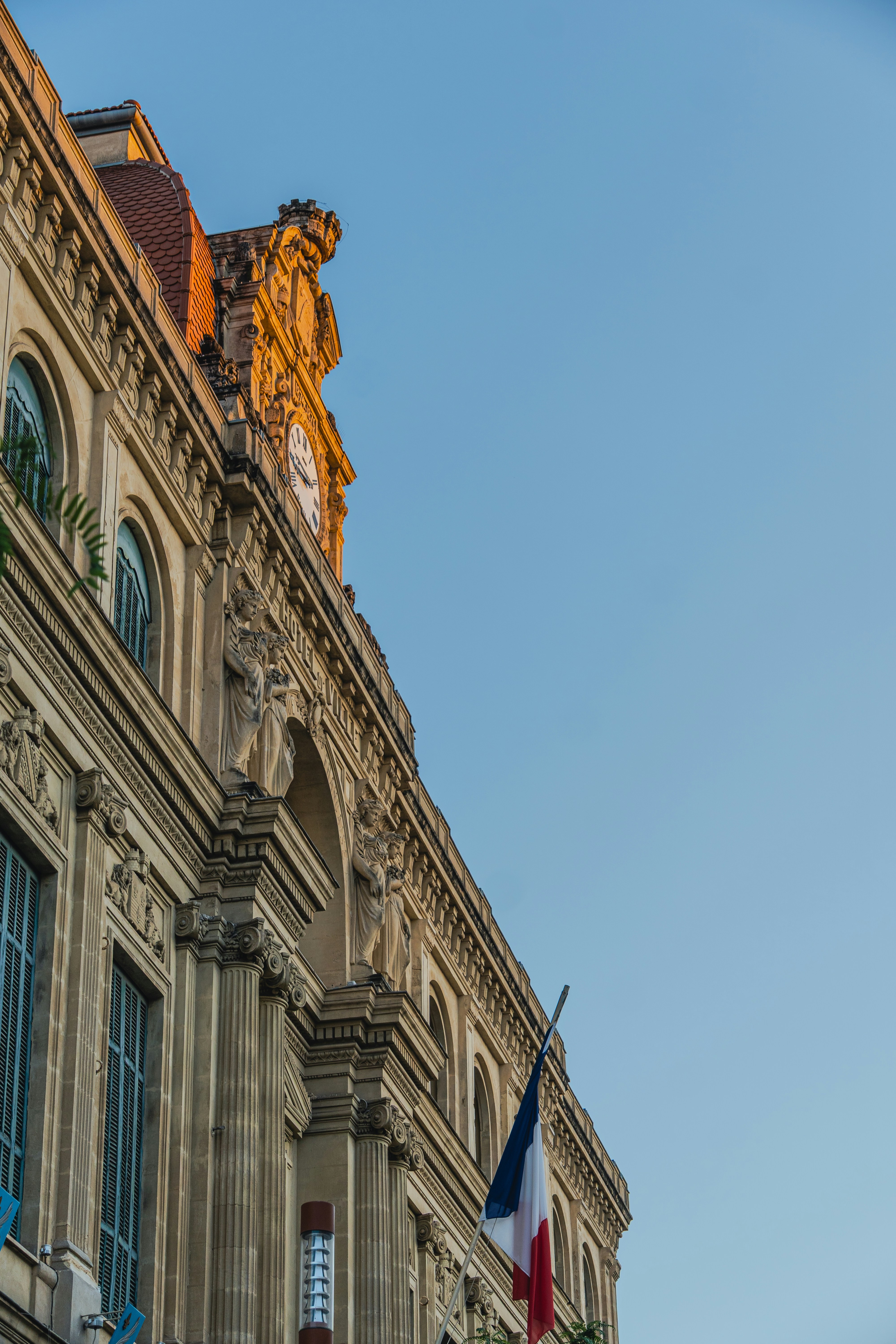 Historic building adorned with intricate details and a clock tower, illuminated by the warm glow of sunset. French flag gently waving in the foreground.