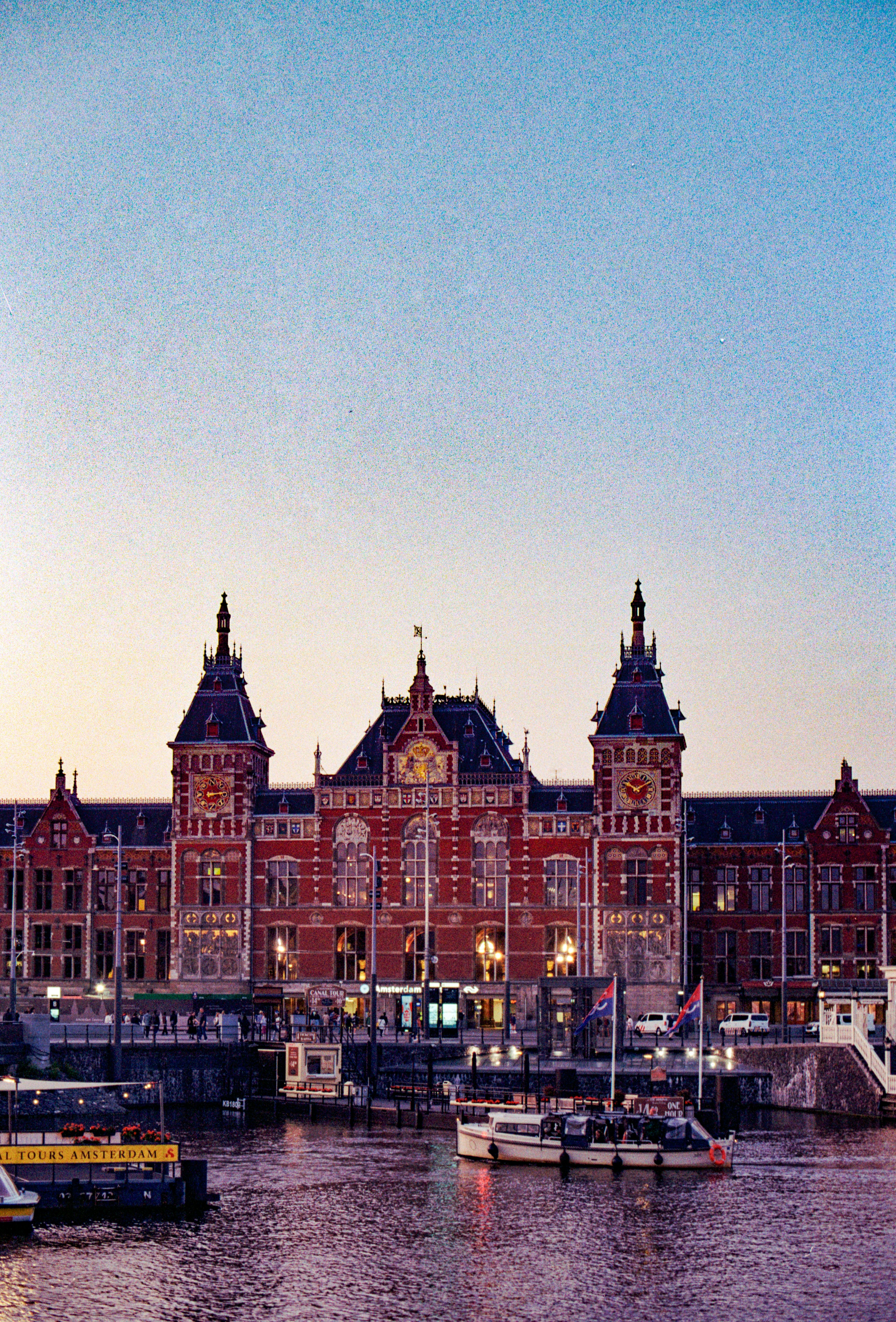 Historic train station in Amsterdam illuminated at dusk, showcasing intricate architectural details and vibrant reflections on the water.