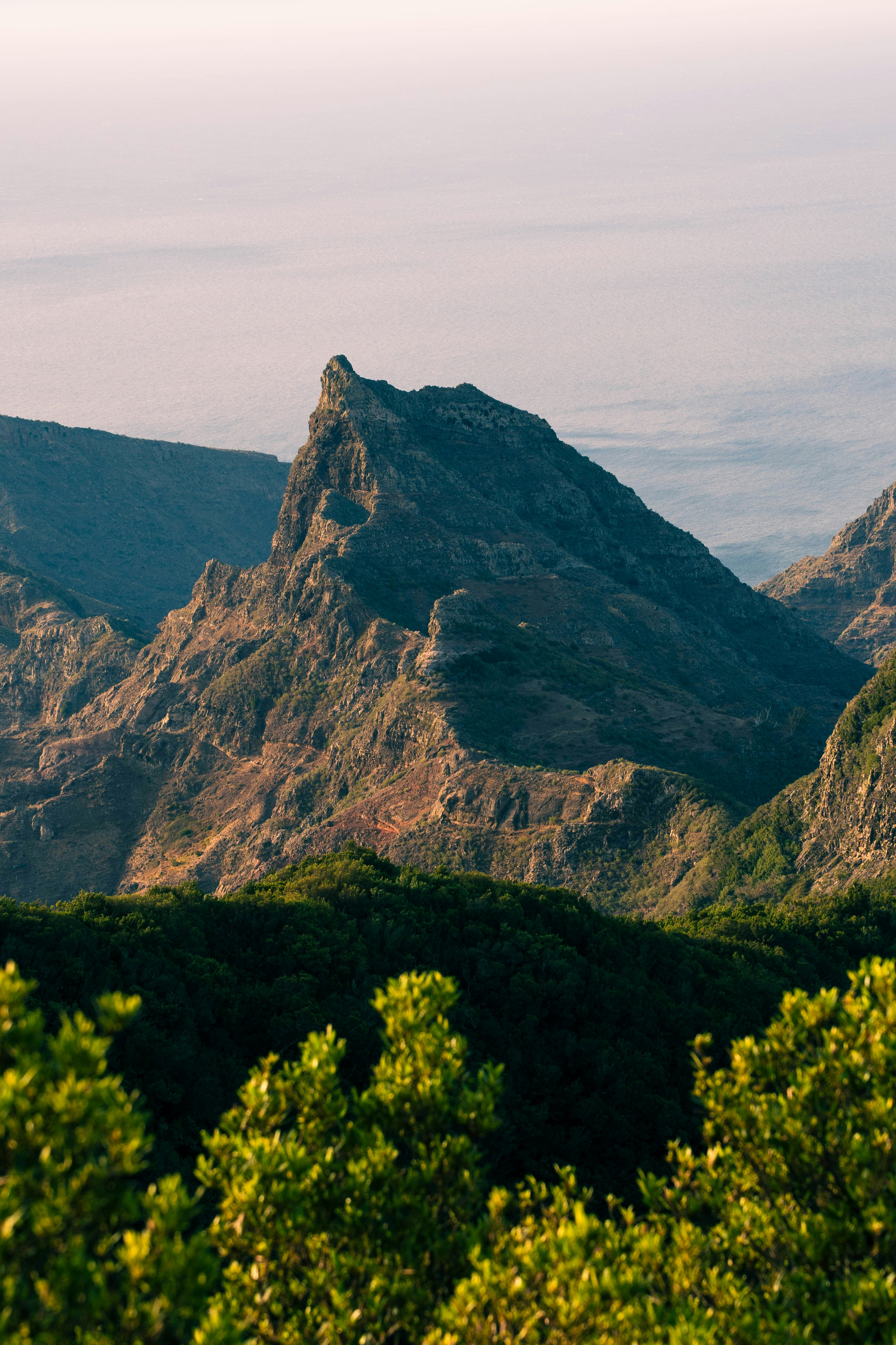 Jagged mountain peak rises majestically above lush green foliage, overlooking a serene ocean expanse. The interplay of light and shadow highlights the rugged terrain.