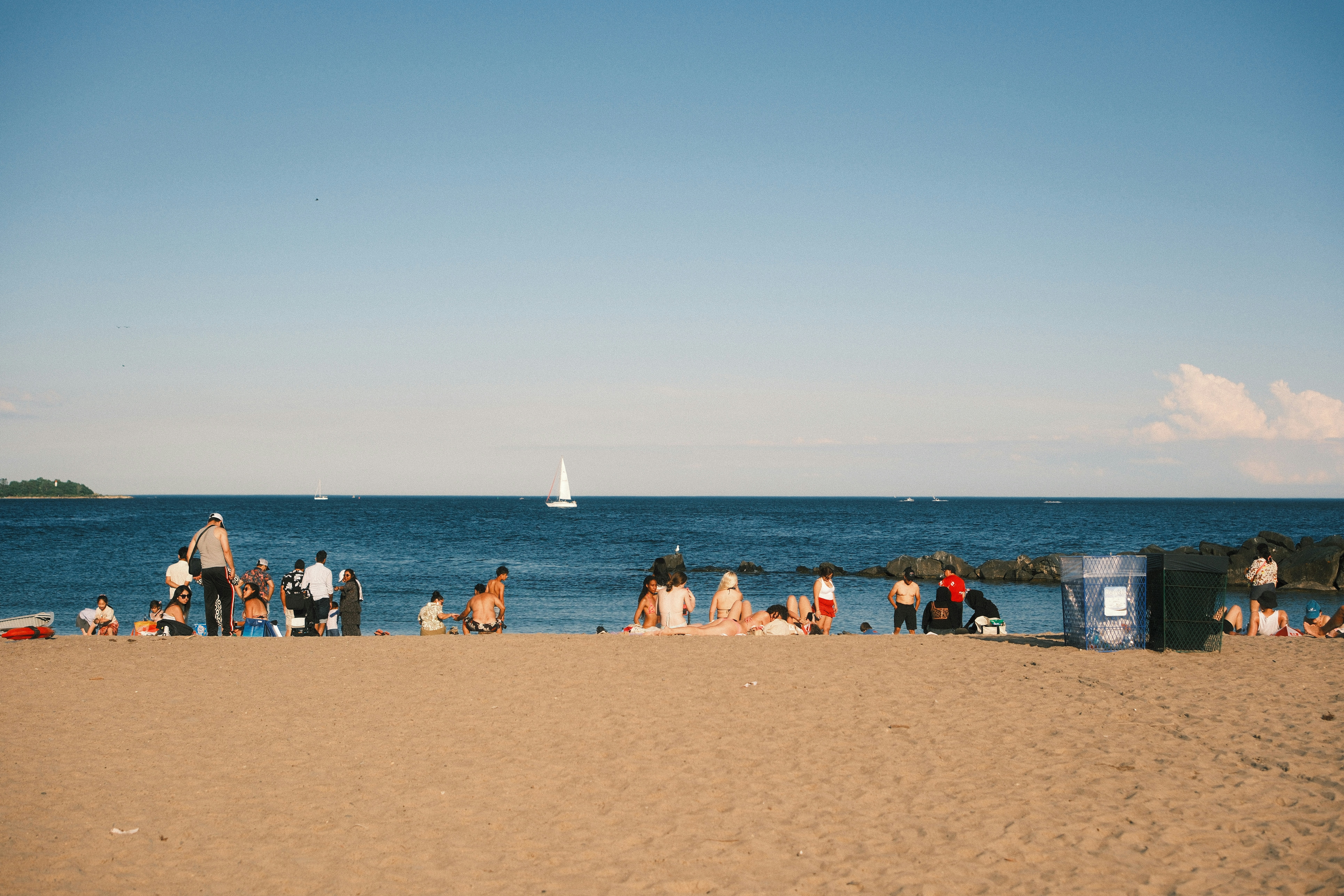 Beachgoers enjoying a sunny day by the water, with a sailboat gliding in the distance. The sandy shore creates a relaxed atmosphere.