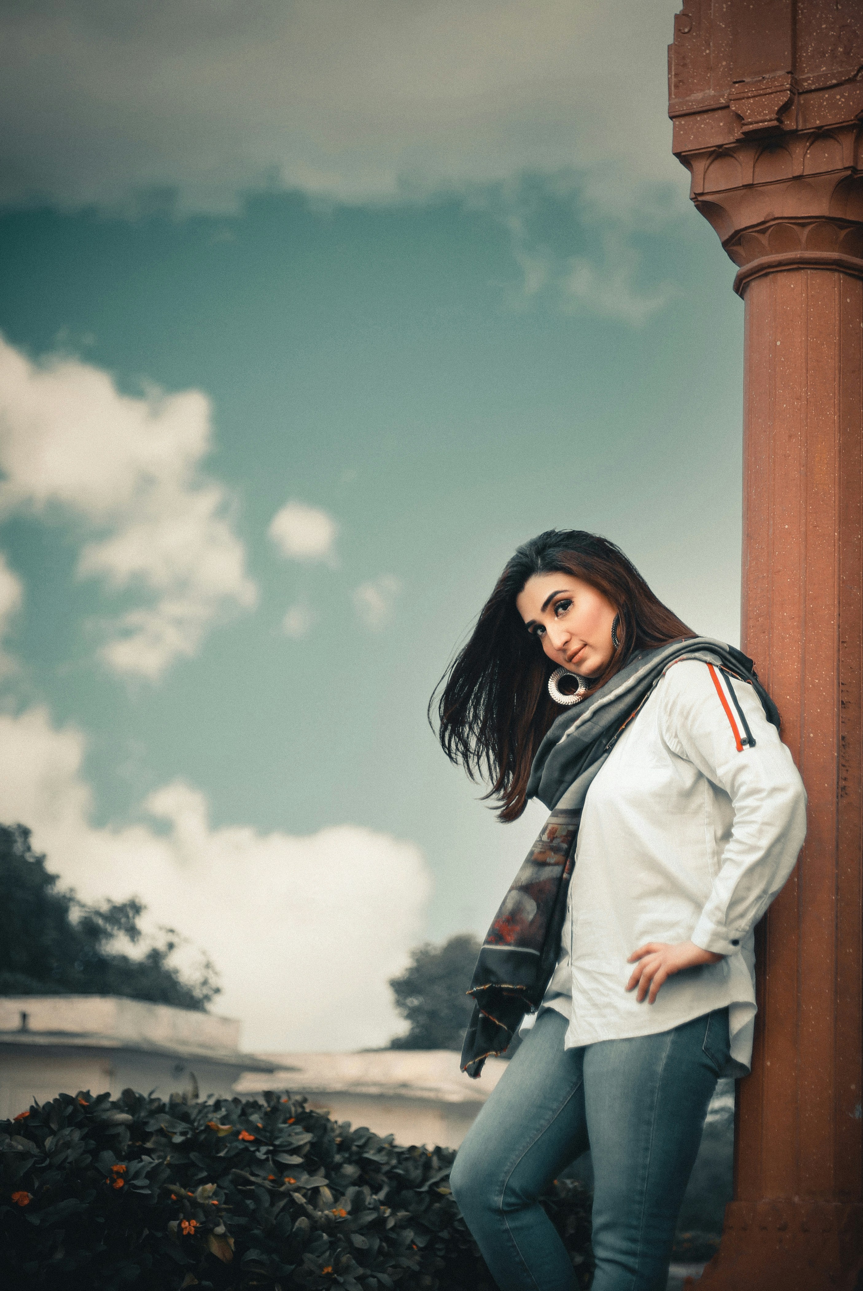 A woman in a stylish outfit leans against a traditional architectural pillar, framed by a vibrant sky and lush greenery.