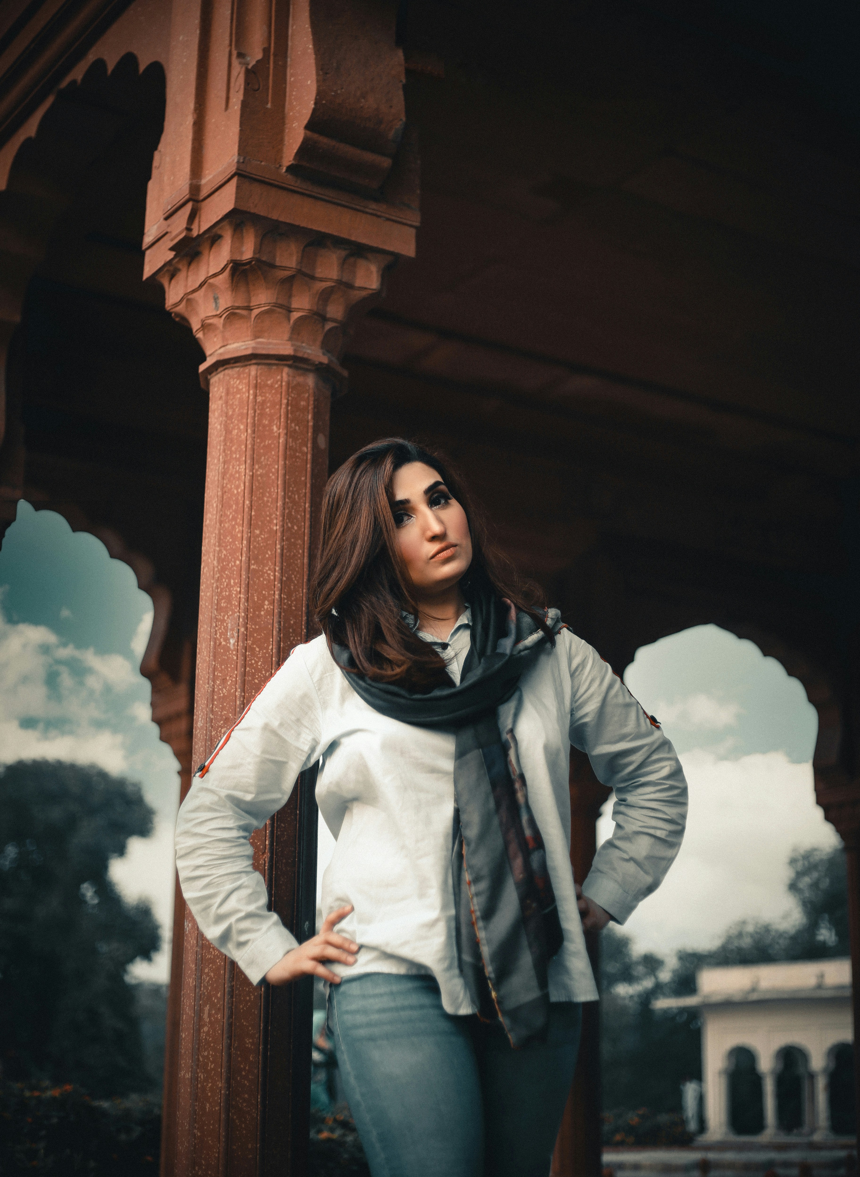 A woman poses thoughtfully against intricately carved pillars, showcasing a blend of modern attire and historic surroundings.