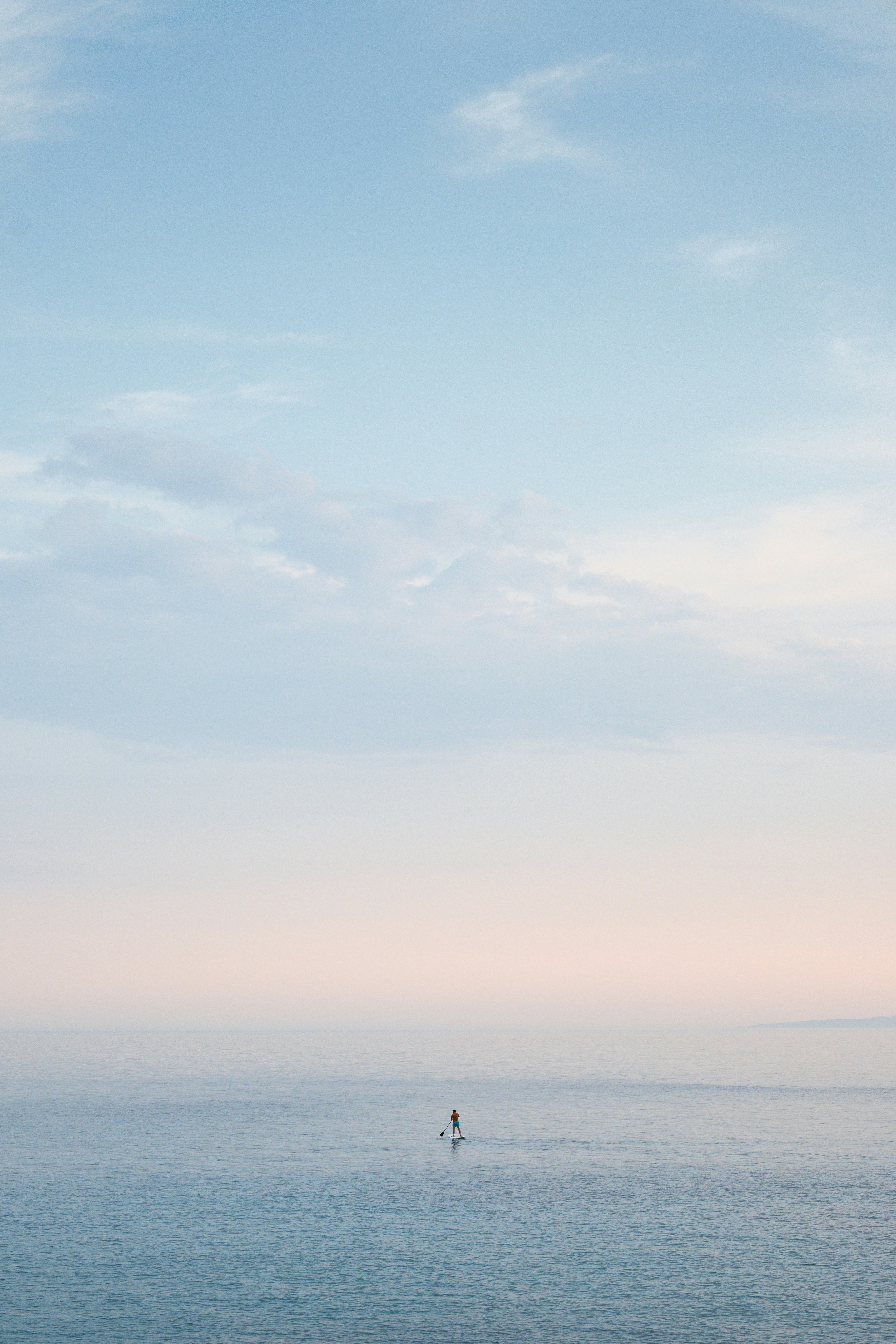 A lone paddleboarder glides across calm sea waters under a pastel sky, capturing a peaceful and minimalist moment at sunrise or sunset. | A person paddleboards on a calm ocean.