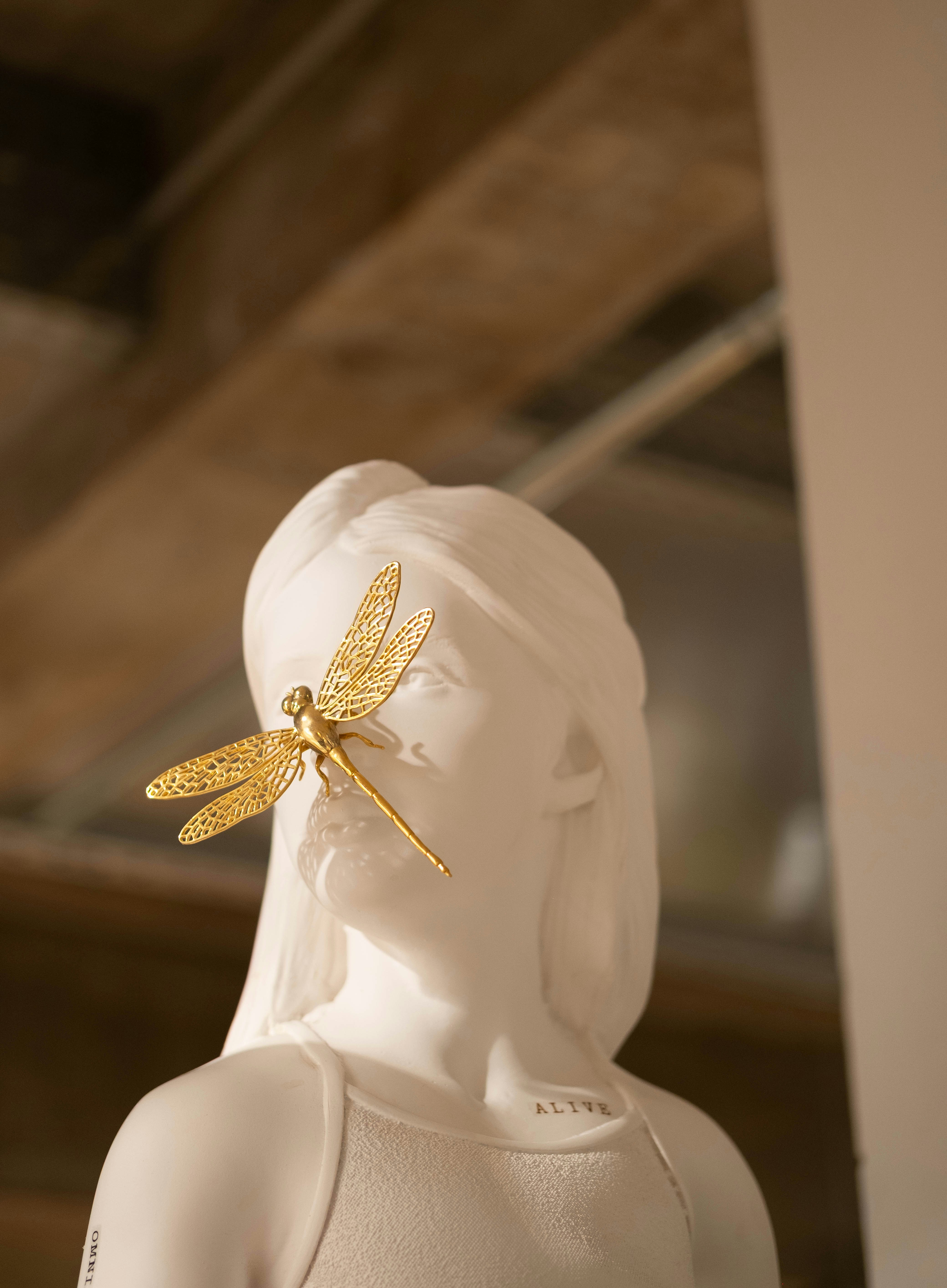 A white sculpture of a woman with a golden dragonfly perched on her face, symbolizing a connection between art and nature.