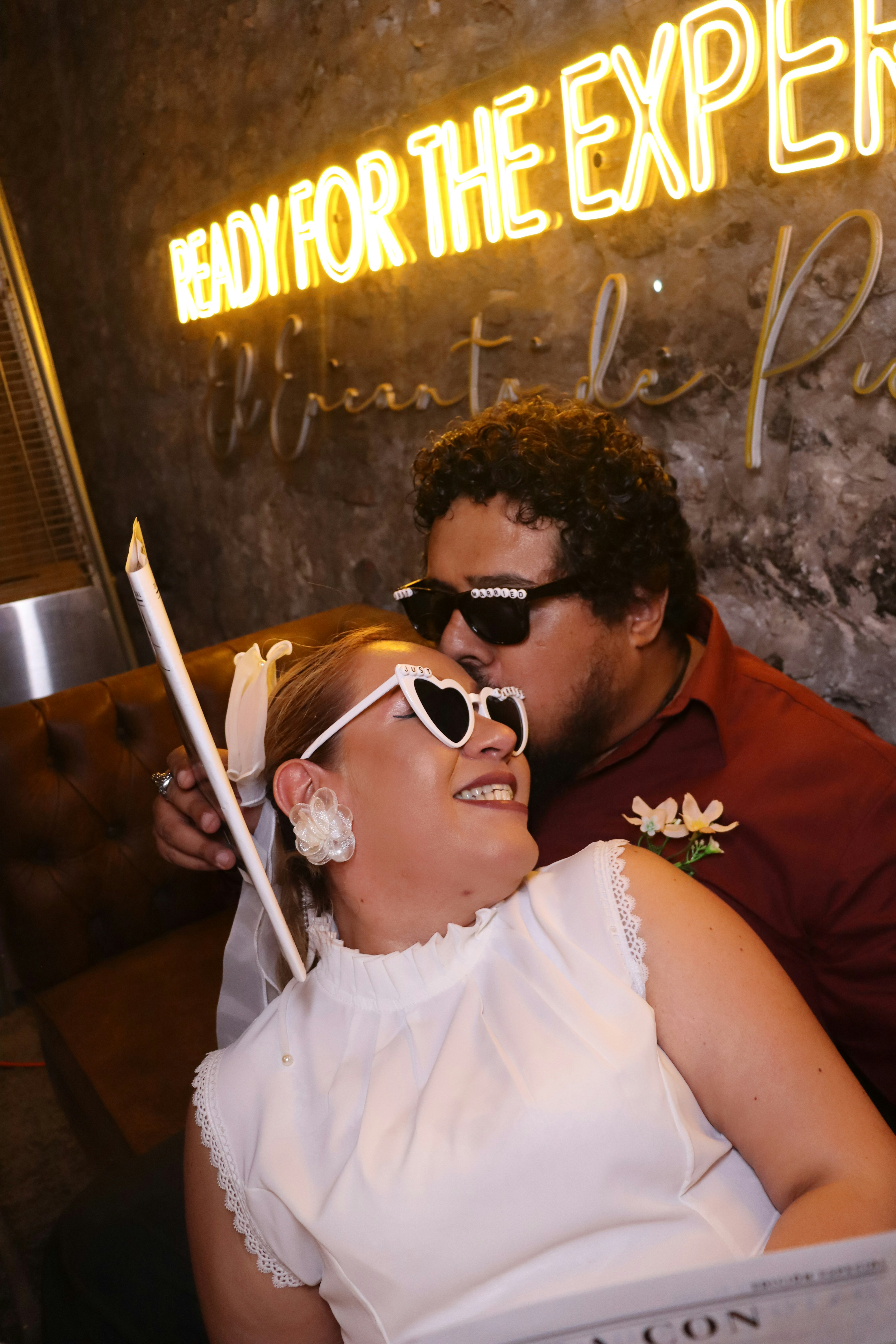 Couple sharing a playful moment in a cozy setting, illuminated by a neon sign. The bride wears heart-shaped sunglasses, adding a whimsical touch.