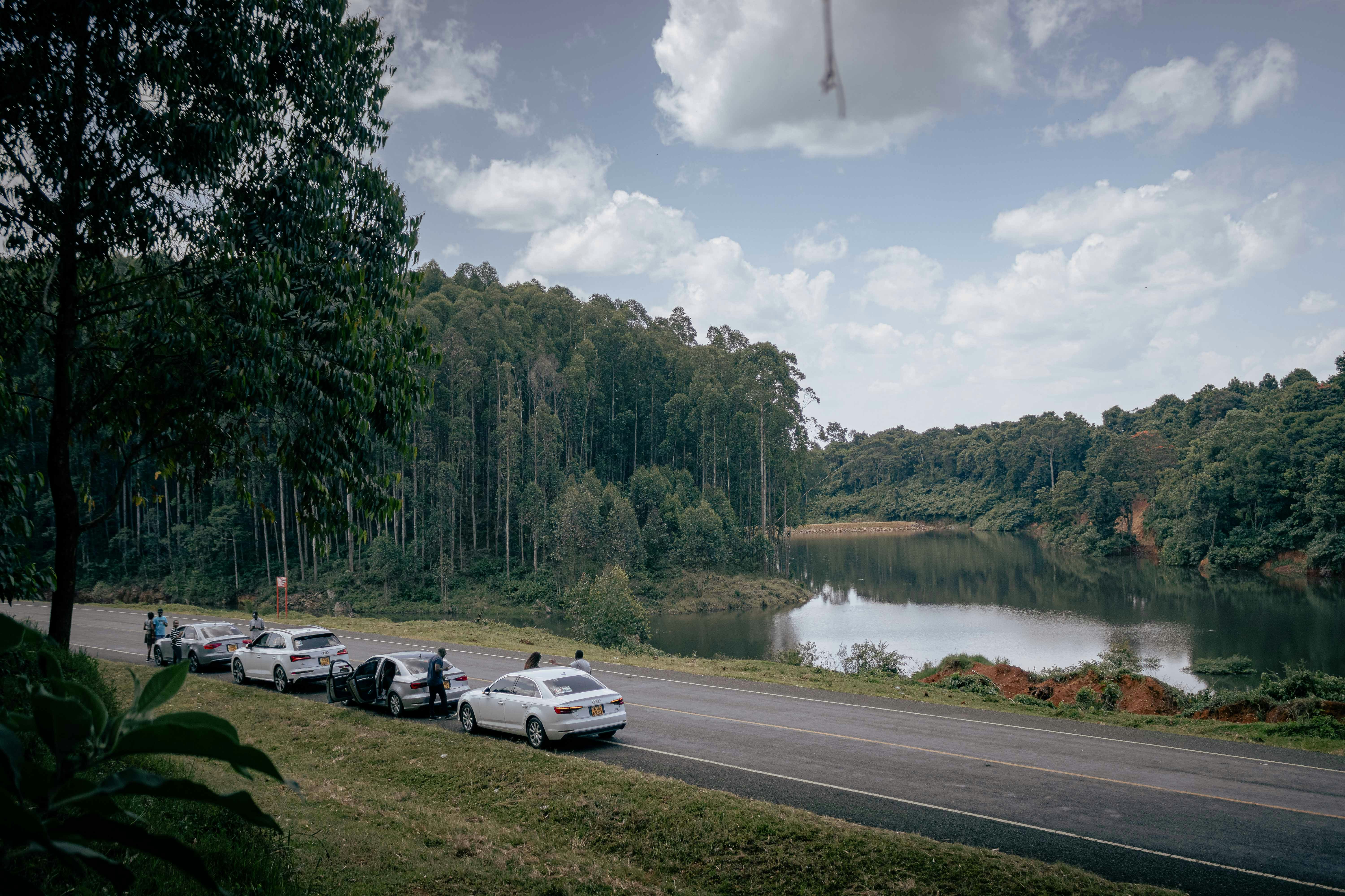 Cars parked along a winding road near a tranquil lake surrounded by lush greenery and distant hills. A few people engage in conversation, enjoying the peaceful atmosphere.