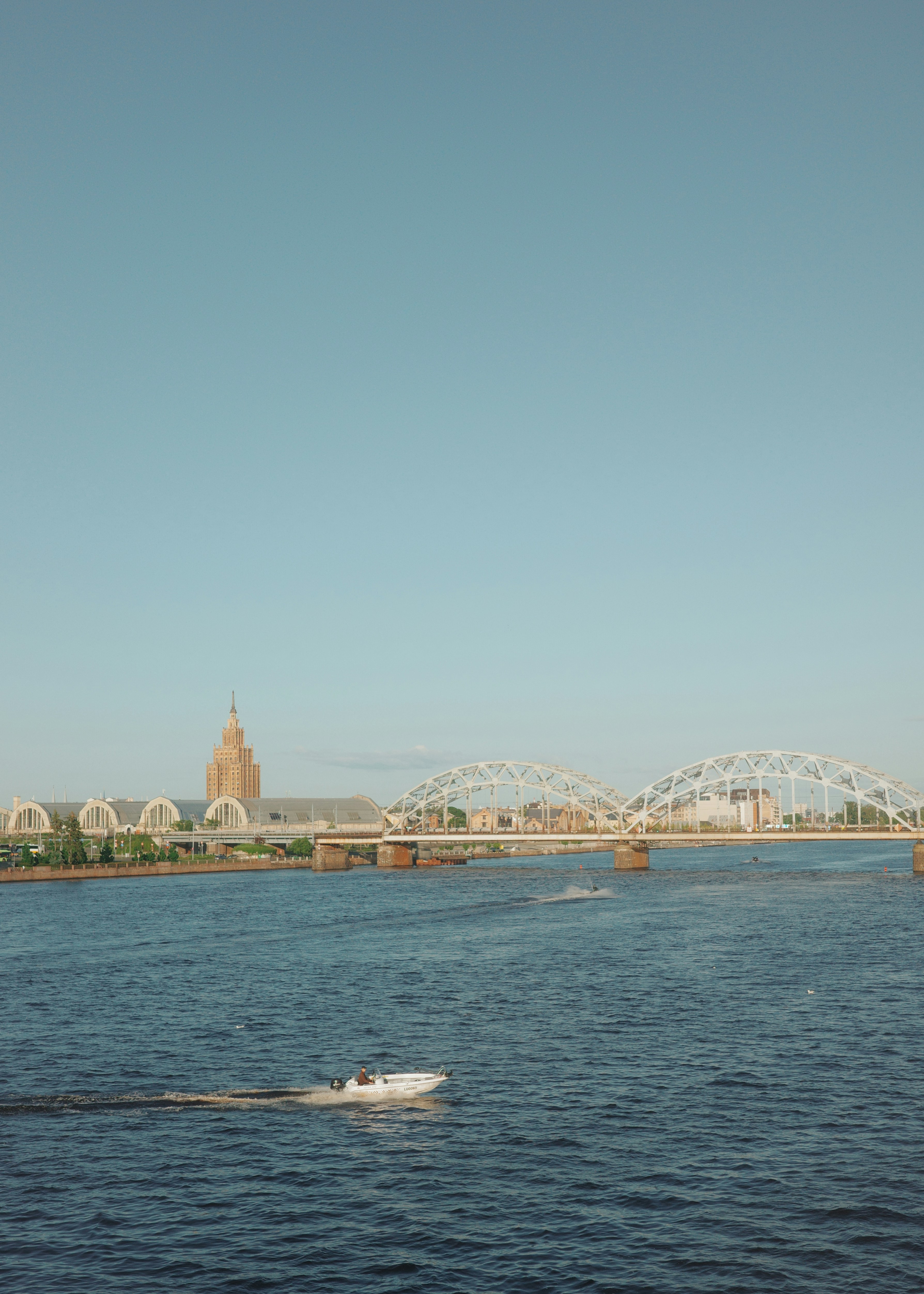 A boat glides across the river beneath architectural bridges, with a prominent city skyline in the background. The scene captures a moment of tranquility in an urban setting.