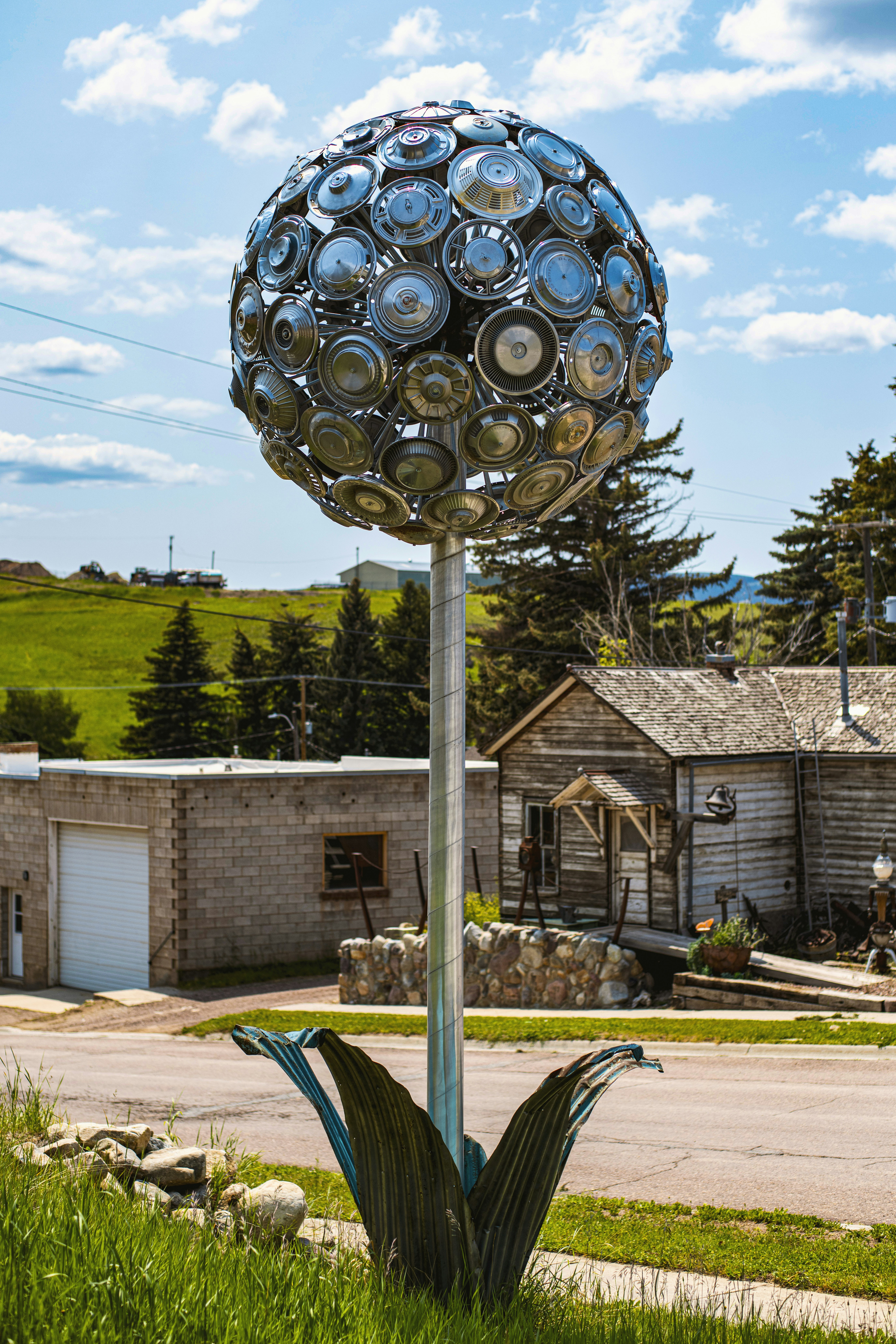 Sculptural flower made from repurposed metal objects, standing prominently against a rural backdrop. The artwork showcases creativity and environmental consciousness.