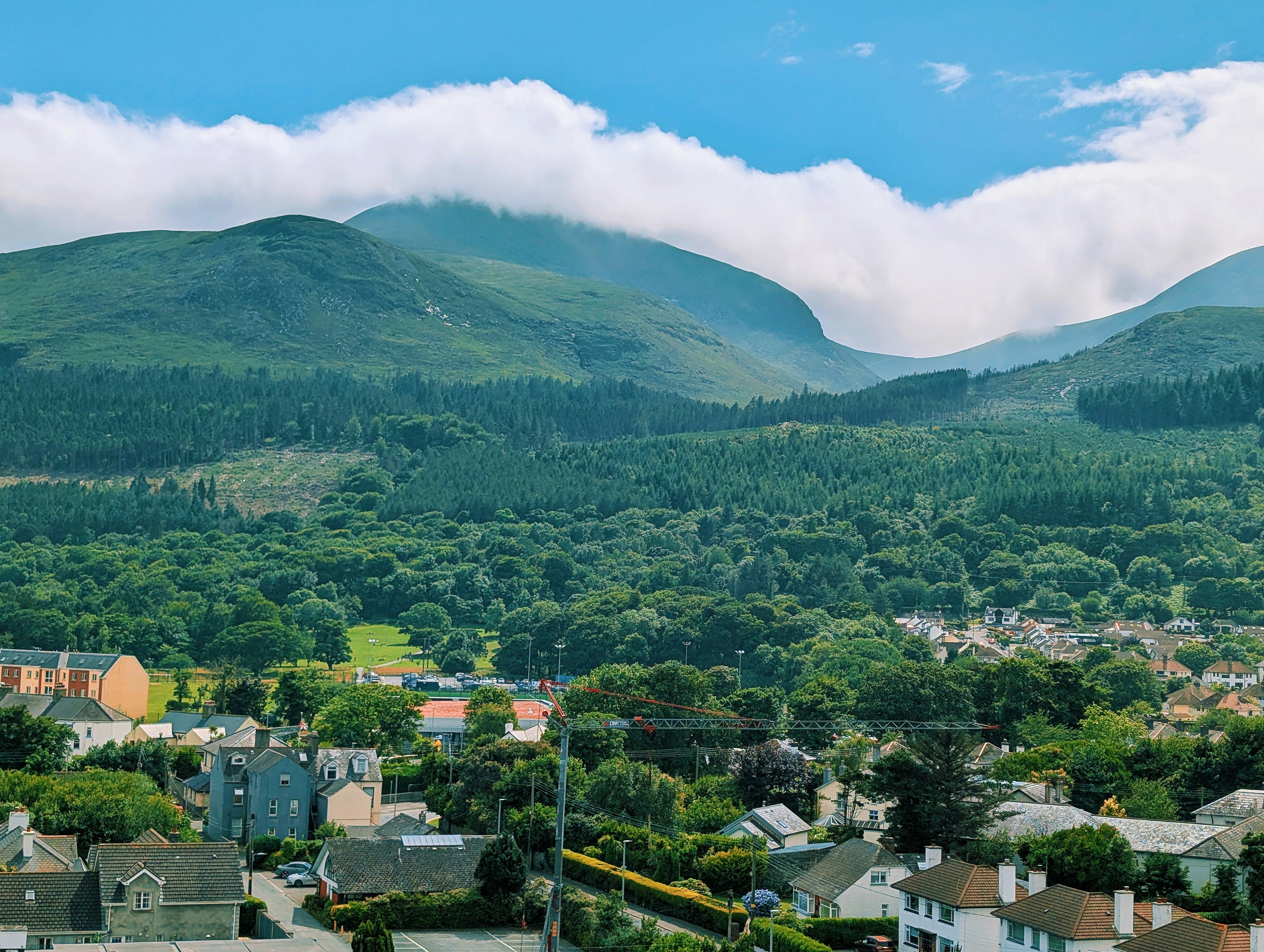 Lush green hills of the Mourne Mountains rise majestically, partially shrouded in clouds, overlooking a quaint town below.