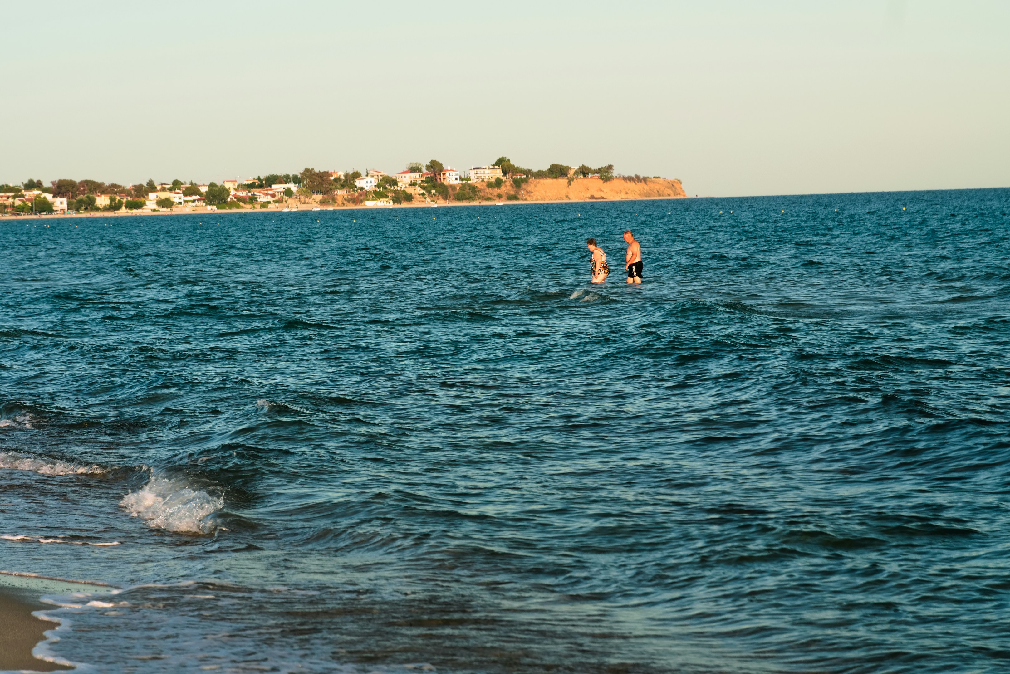 Two figures wade through gentle waves under a soft twilight sky, with a coastal landscape in the background.