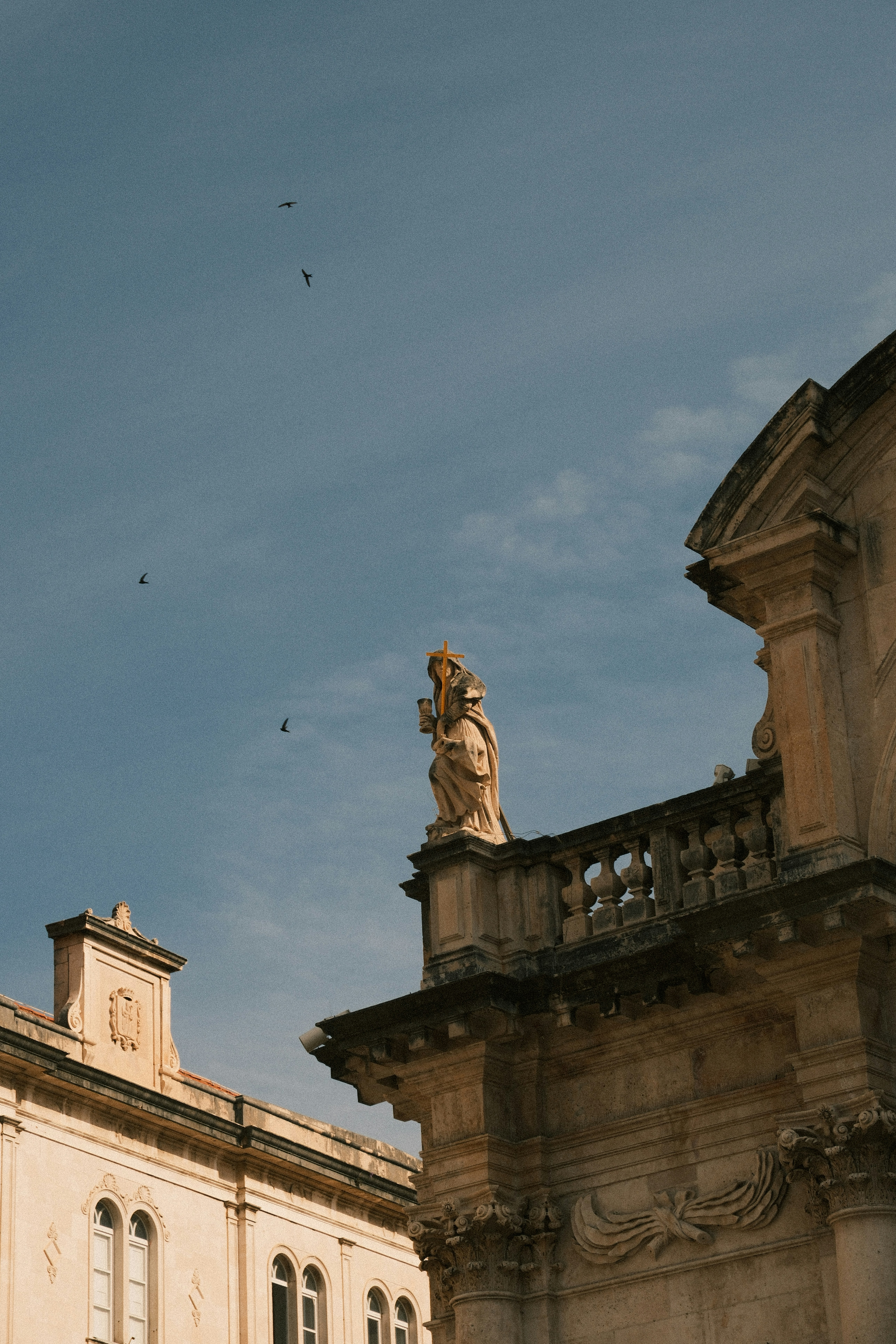 A majestic statue overlooking a historic building under a clear blue sky, with birds soaring in the background.
