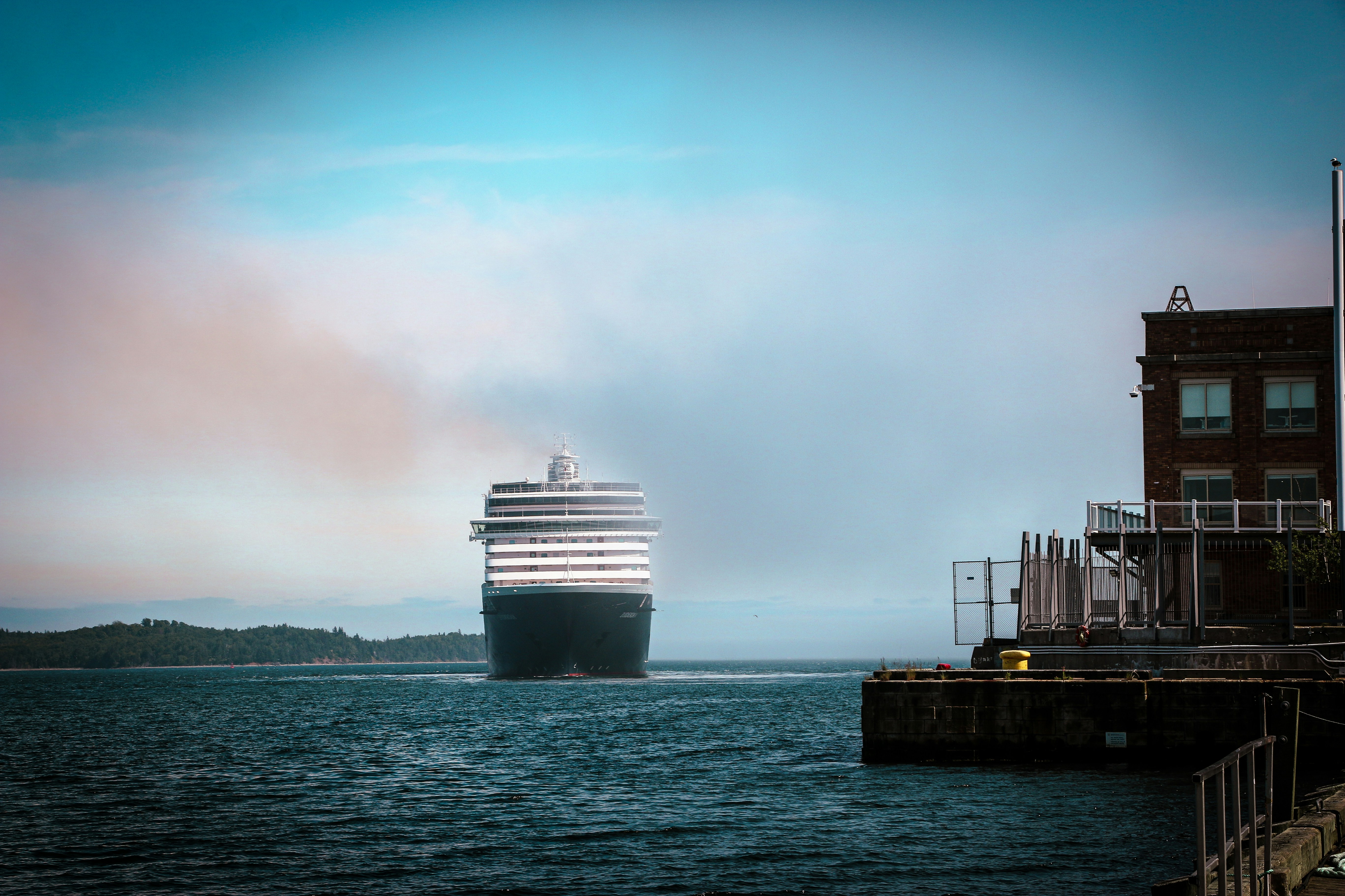 A large cruise ship navigates through a misty harbor, with a backdrop of distant hills and a historic building on the shore.