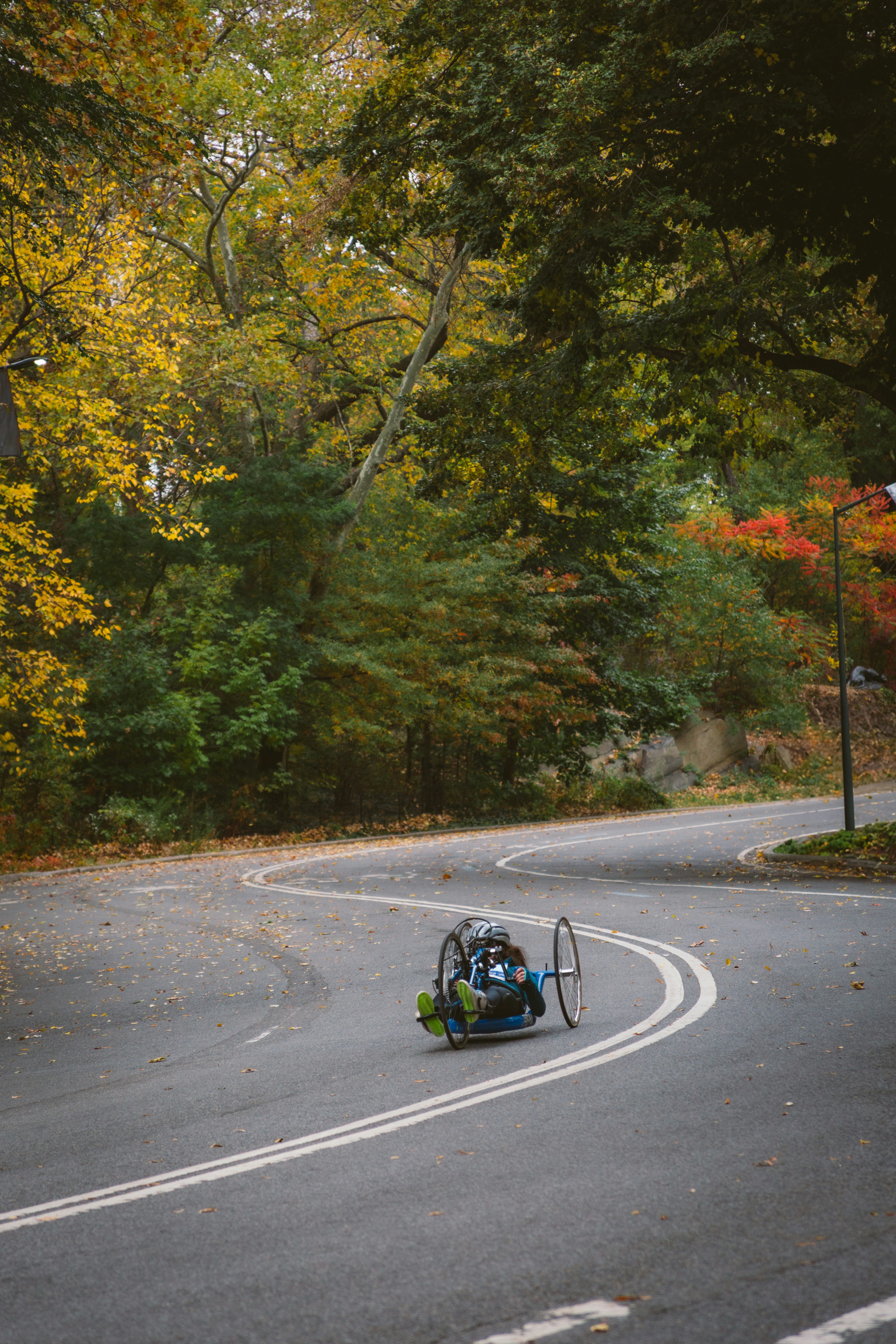 A person maneuvering a handcycle along a curving road surrounded by vibrant autumn foliage.