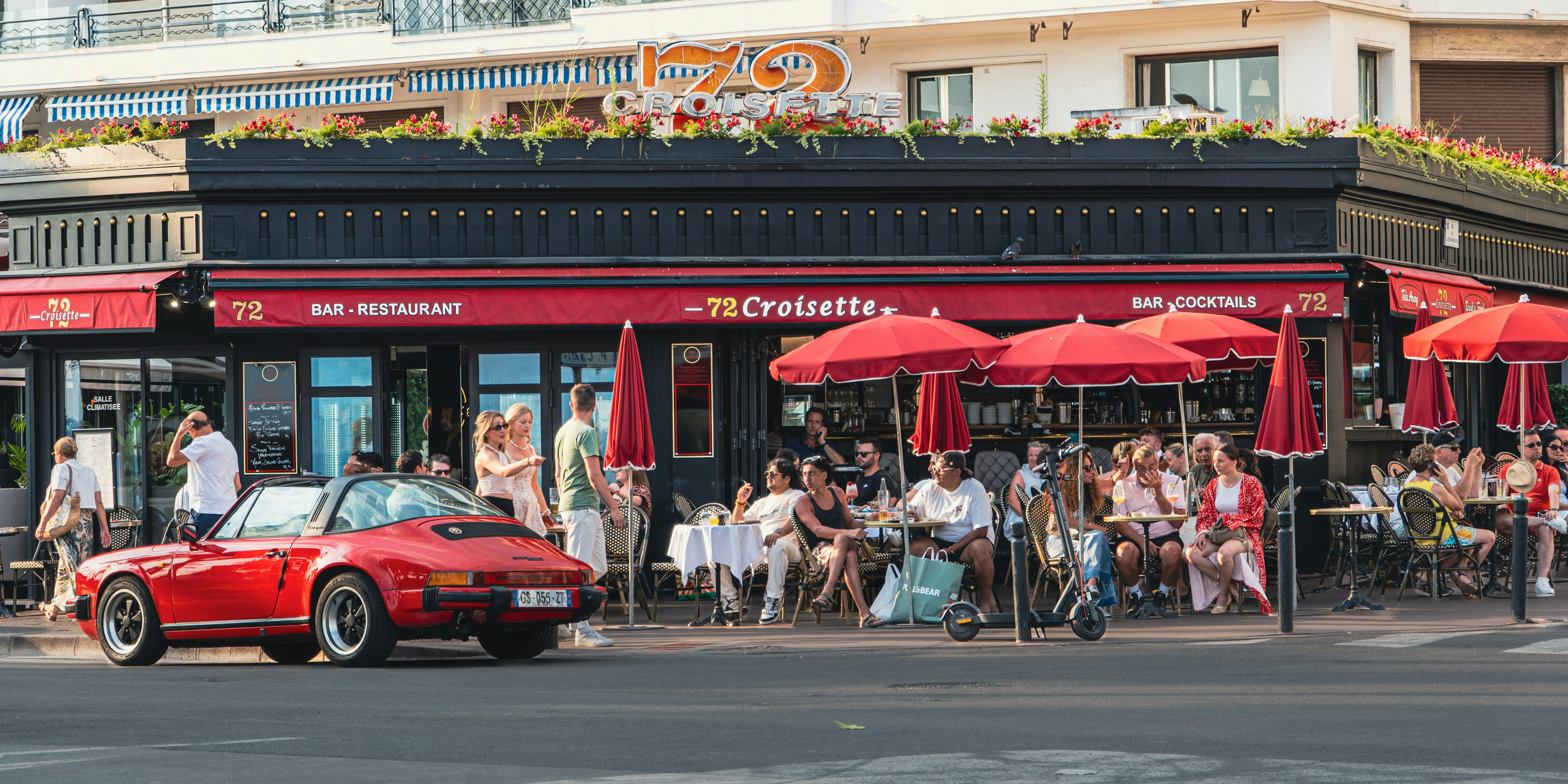 Charming restaurant terrace bustling with patrons enjoying drinks under vibrant red umbrellas, alongside a classic car parked nearby.