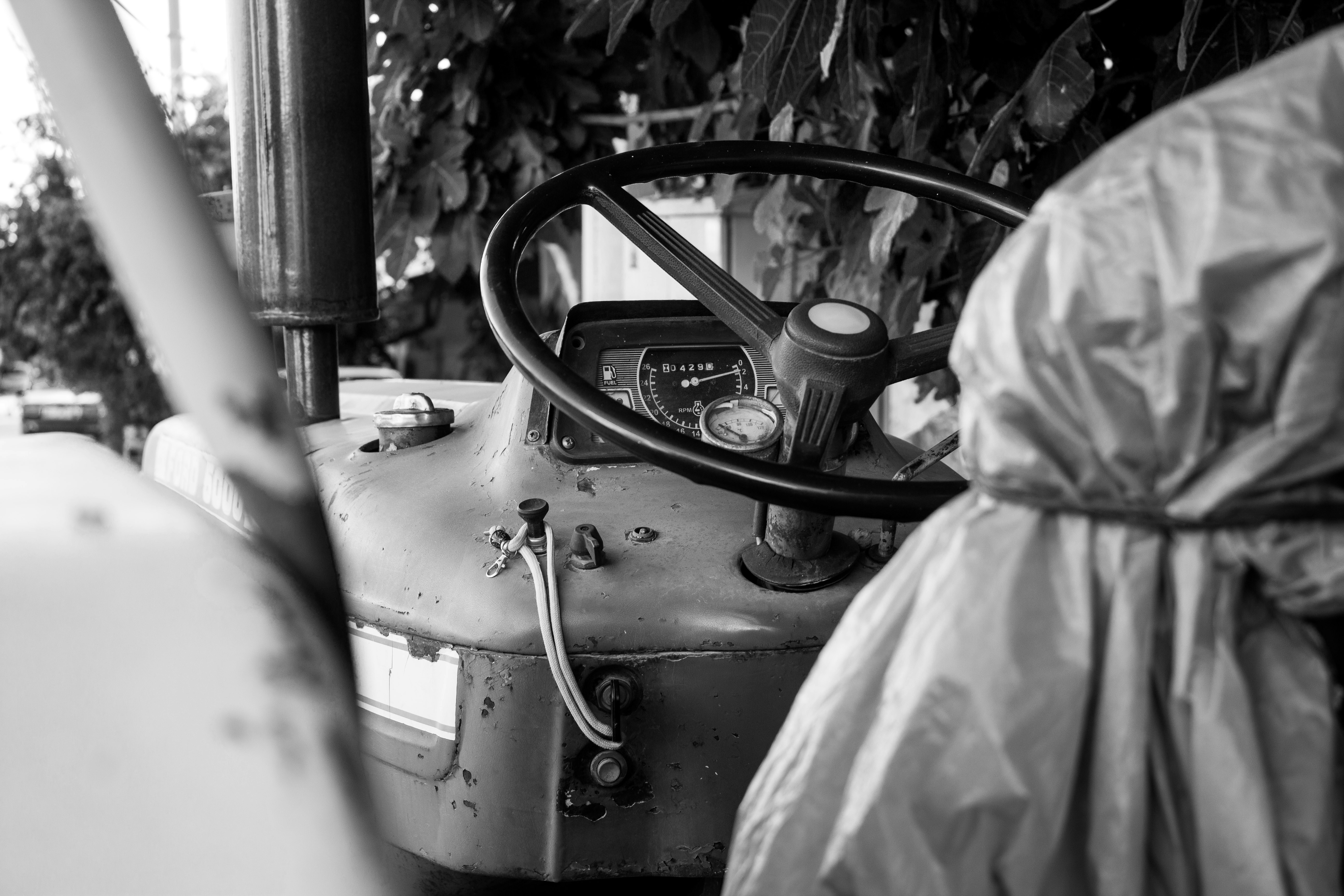Close-up of a tractor's steering wheel and dashboard, showcasing vintage gauges and a tarp draped over the seat. The monochrome tones enhance the nostalgic feel.