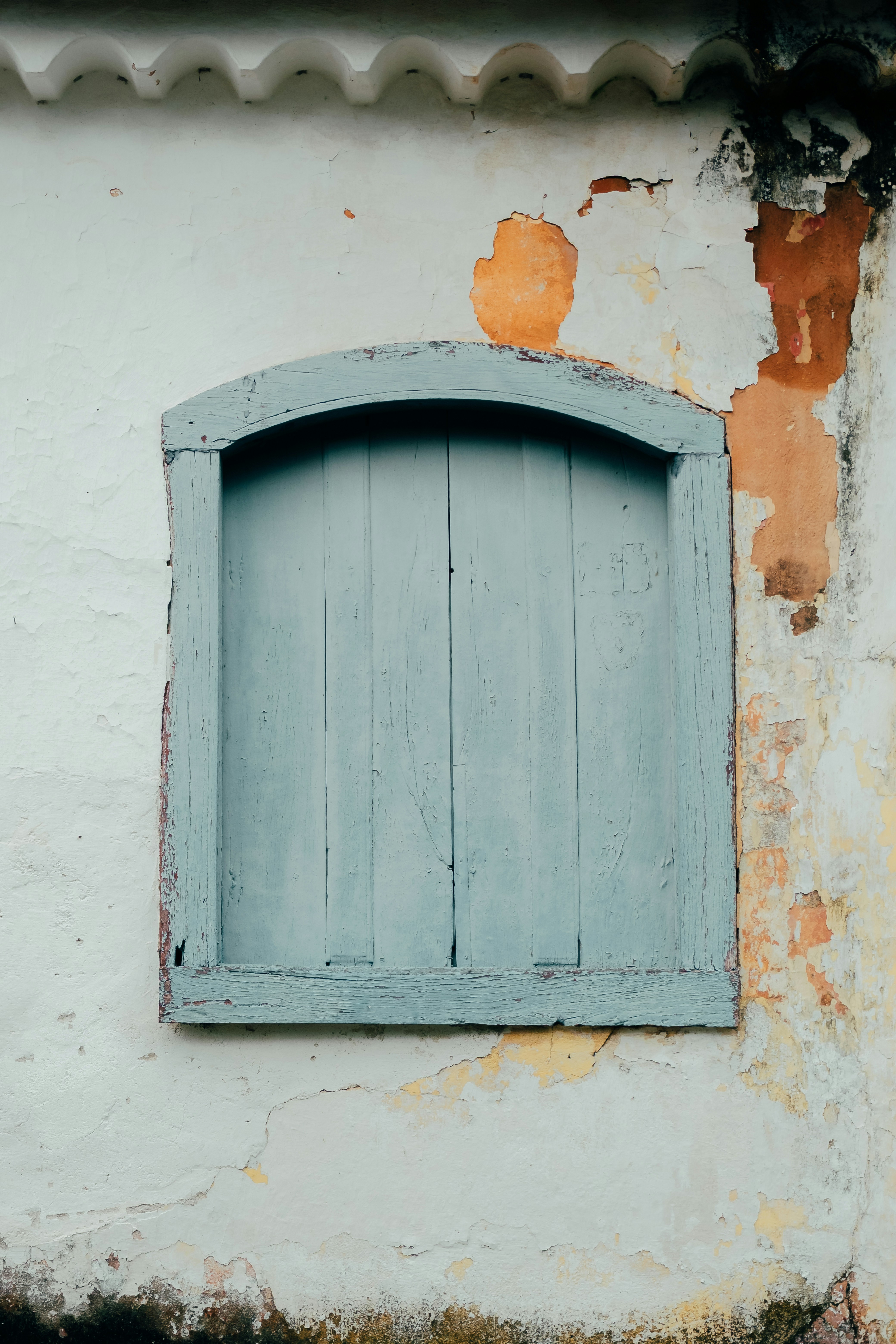 Weathered blue shutter against a peeling white wall, showcasing a blend of colors and textures that tell a story of age and character.