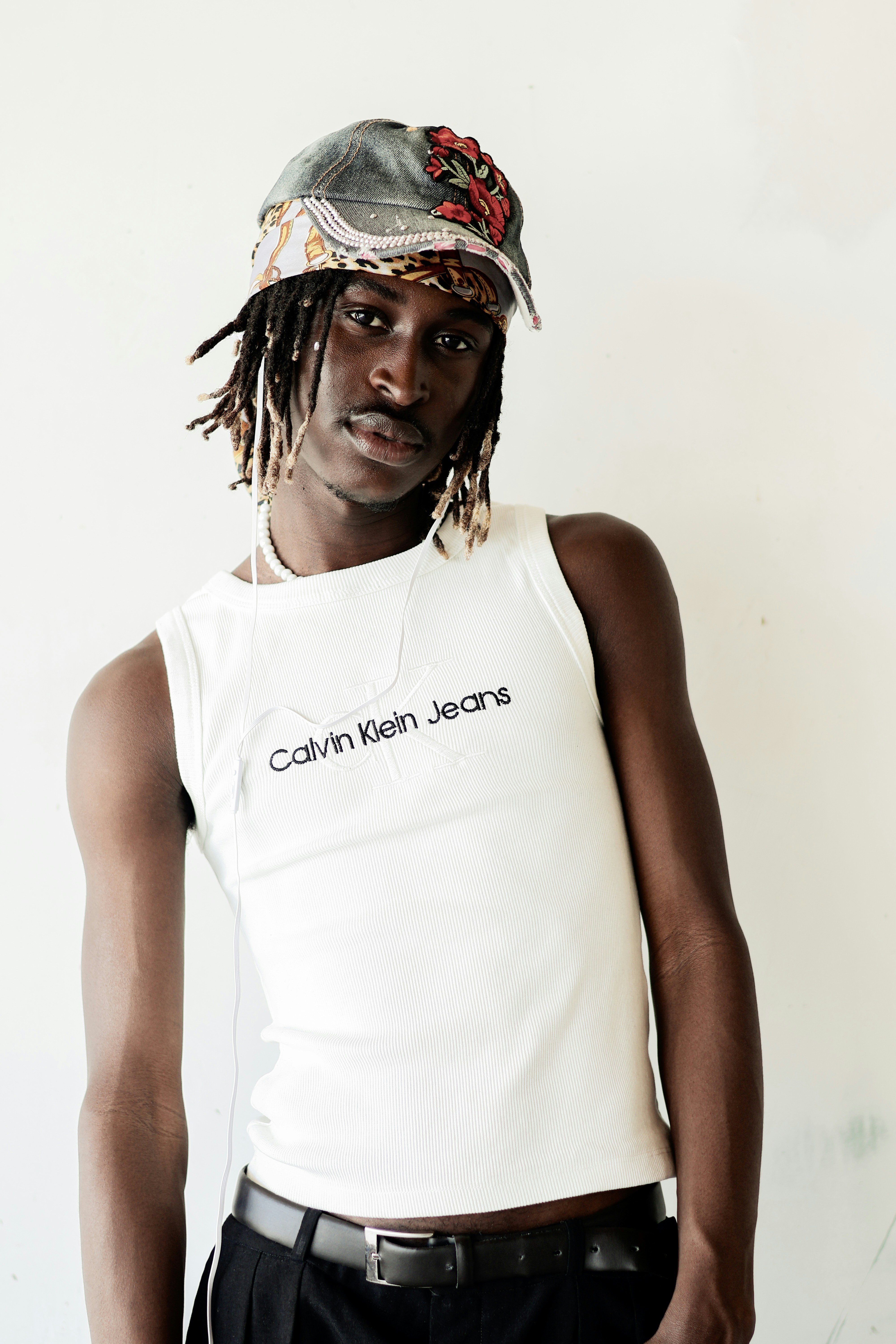 Young man in a white sleeveless top featuring 'Calvin Klein Jeans' logo, complemented by a patterned cap and stylish accessories.