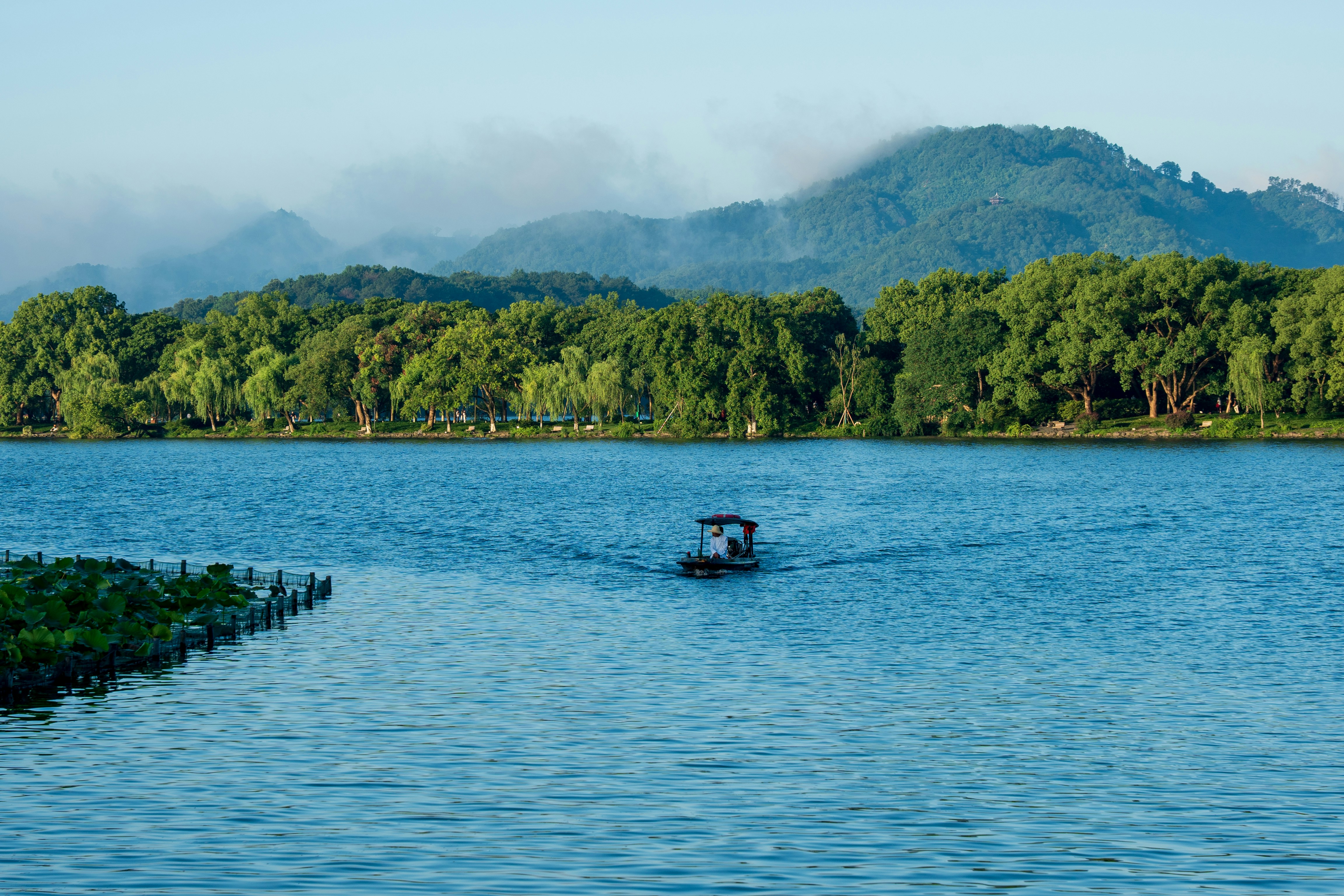 A small boat glides across a tranquil lake surrounded by lush greenery and distant mountains. The calm water reflects the vibrant landscape.