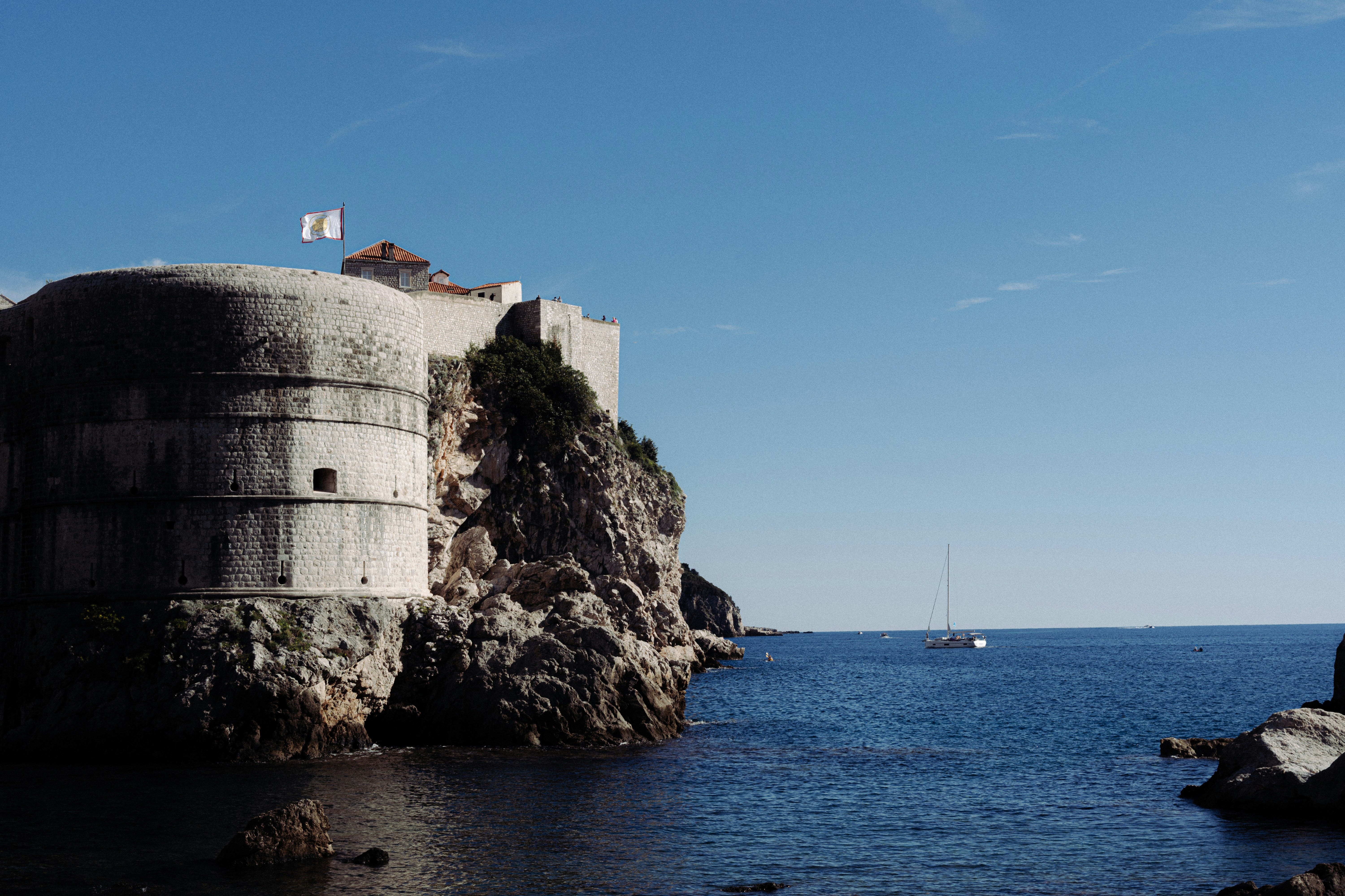 Historic stone fortress perched on a rocky cliff overlooking the calm Adriatic Sea, with a sailboat in the distance. A flag flutters atop the structure.