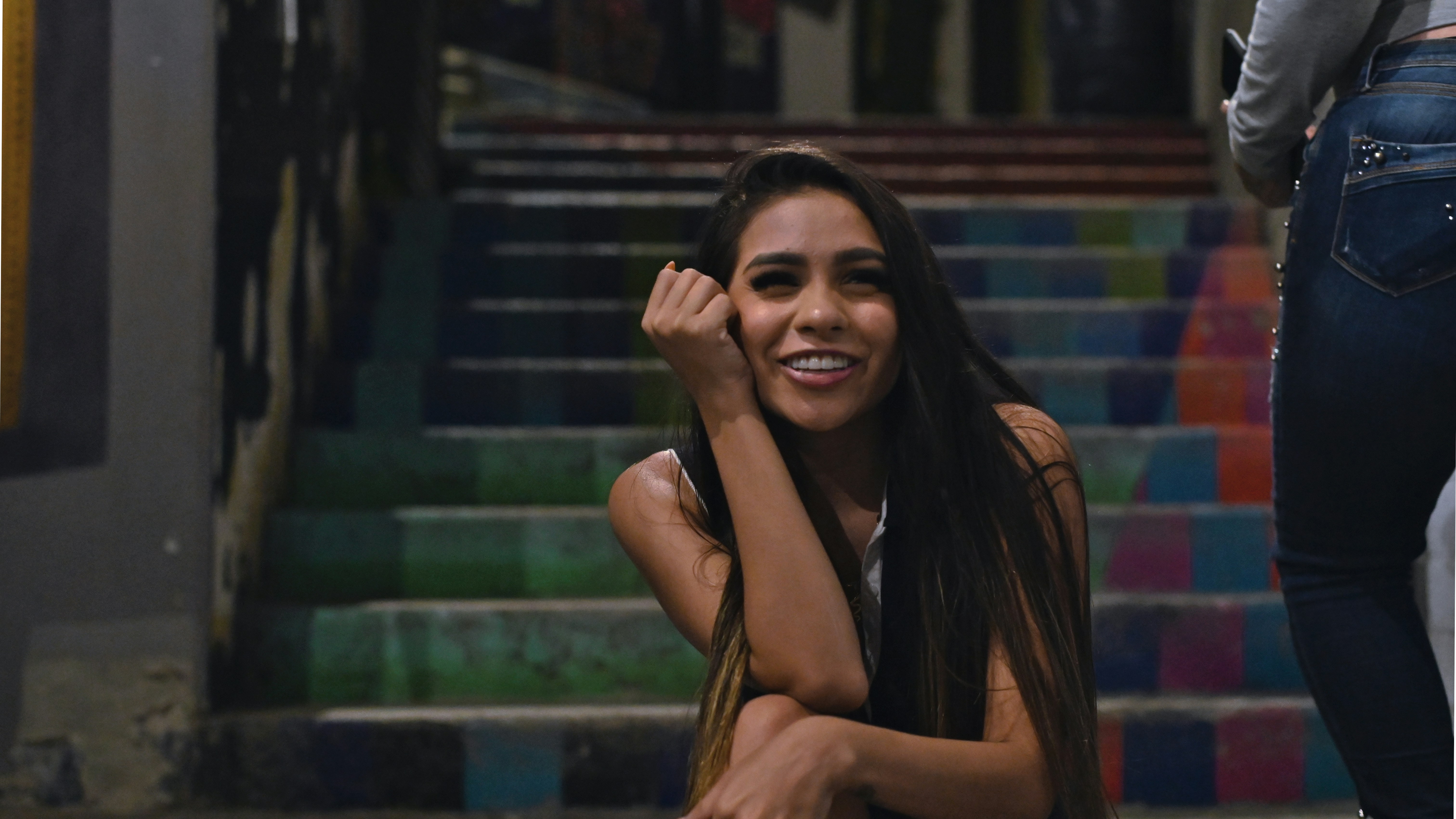 Young woman smiling while sitting on colorful staircase, embodying joy and creativity. The scene captures a lively urban atmosphere.