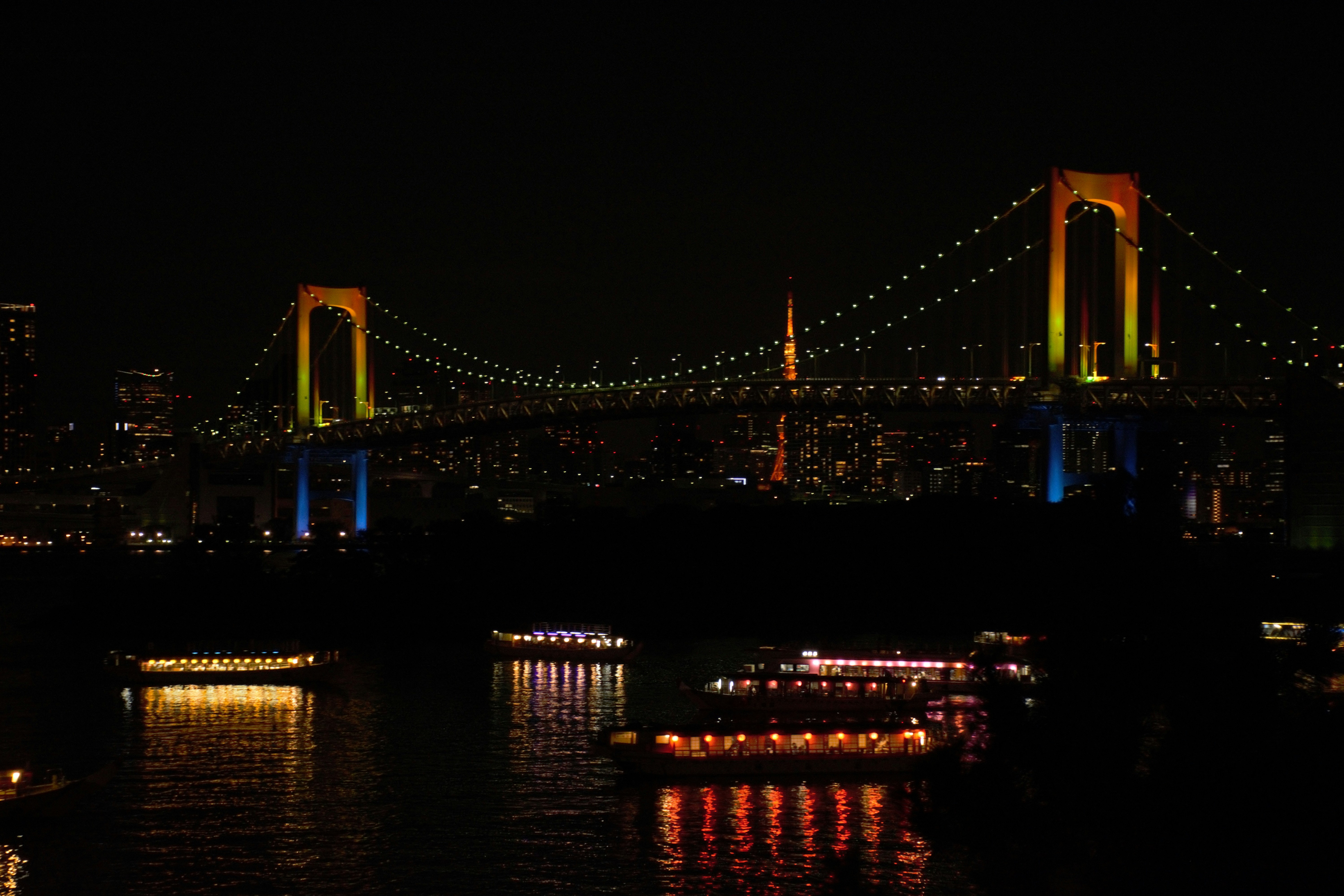 Colorful bridge illuminated against a dark skyline, with boats gliding on the water below.