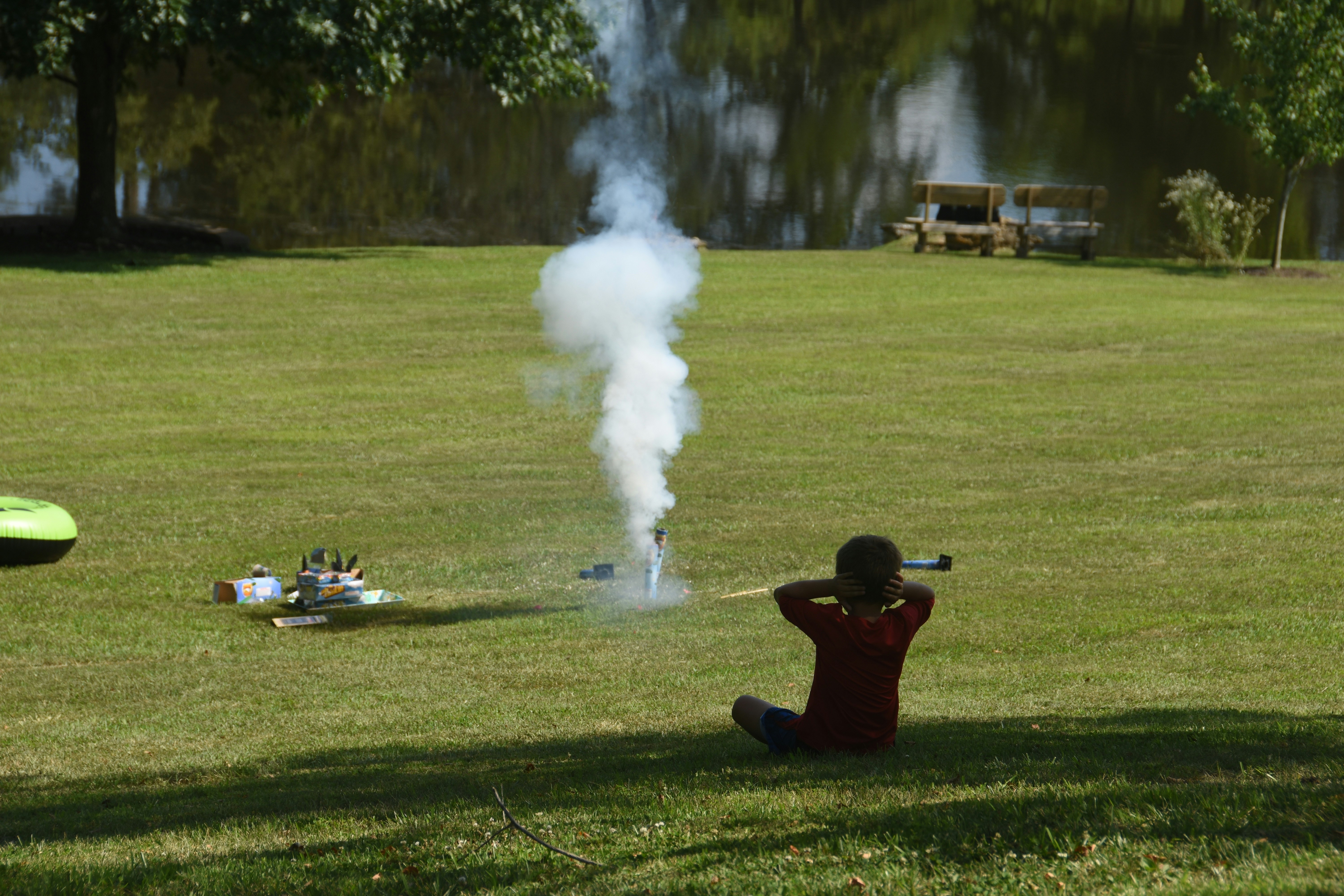 A young child sits with hands over ears as a smoke plume rises from a homemade rocket launch in a grassy field. The serene backdrop features a calm water body and distant seating.