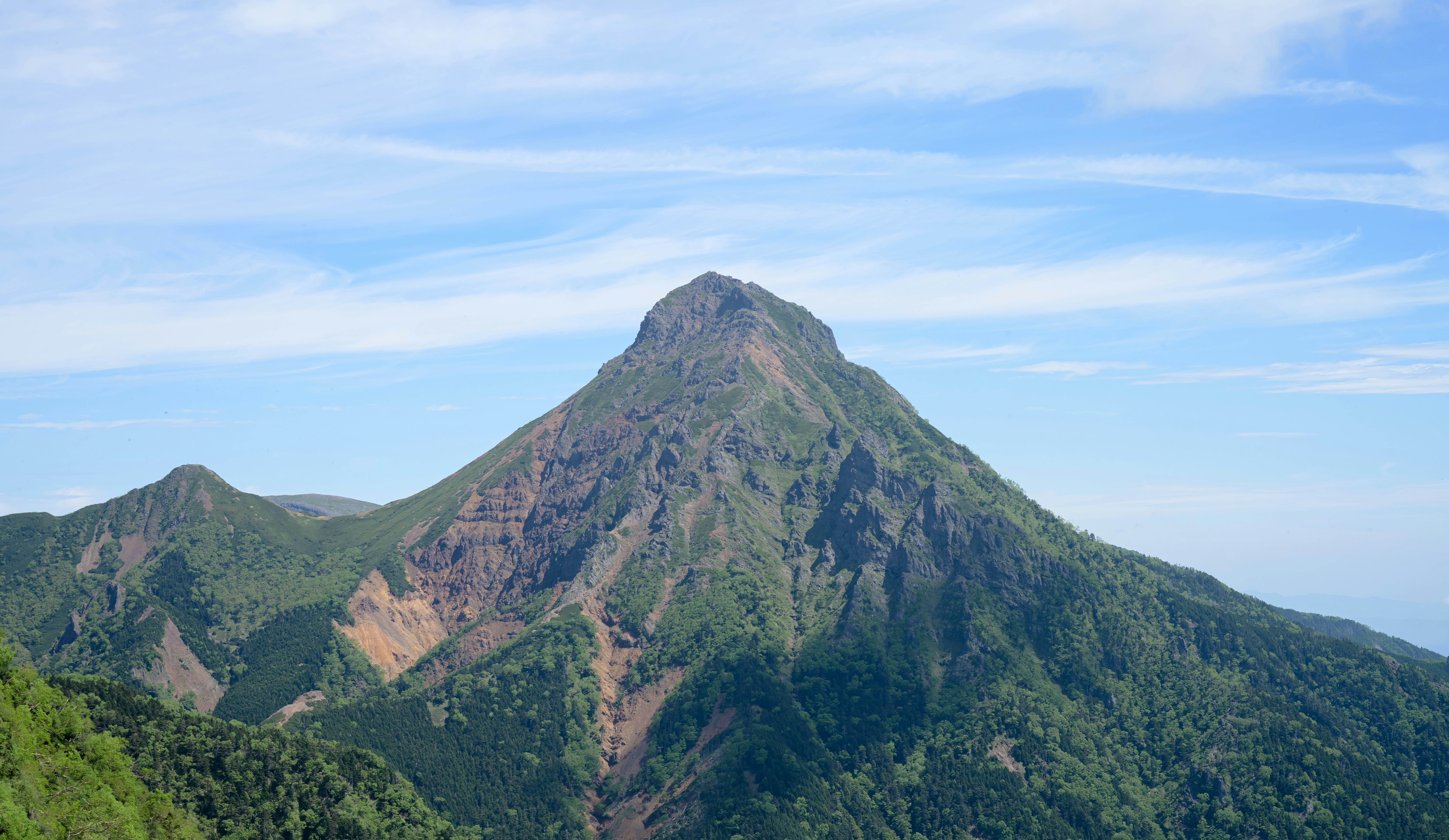 Majestic mountain peak rises above lush green valleys under a blue sky with wispy clouds.