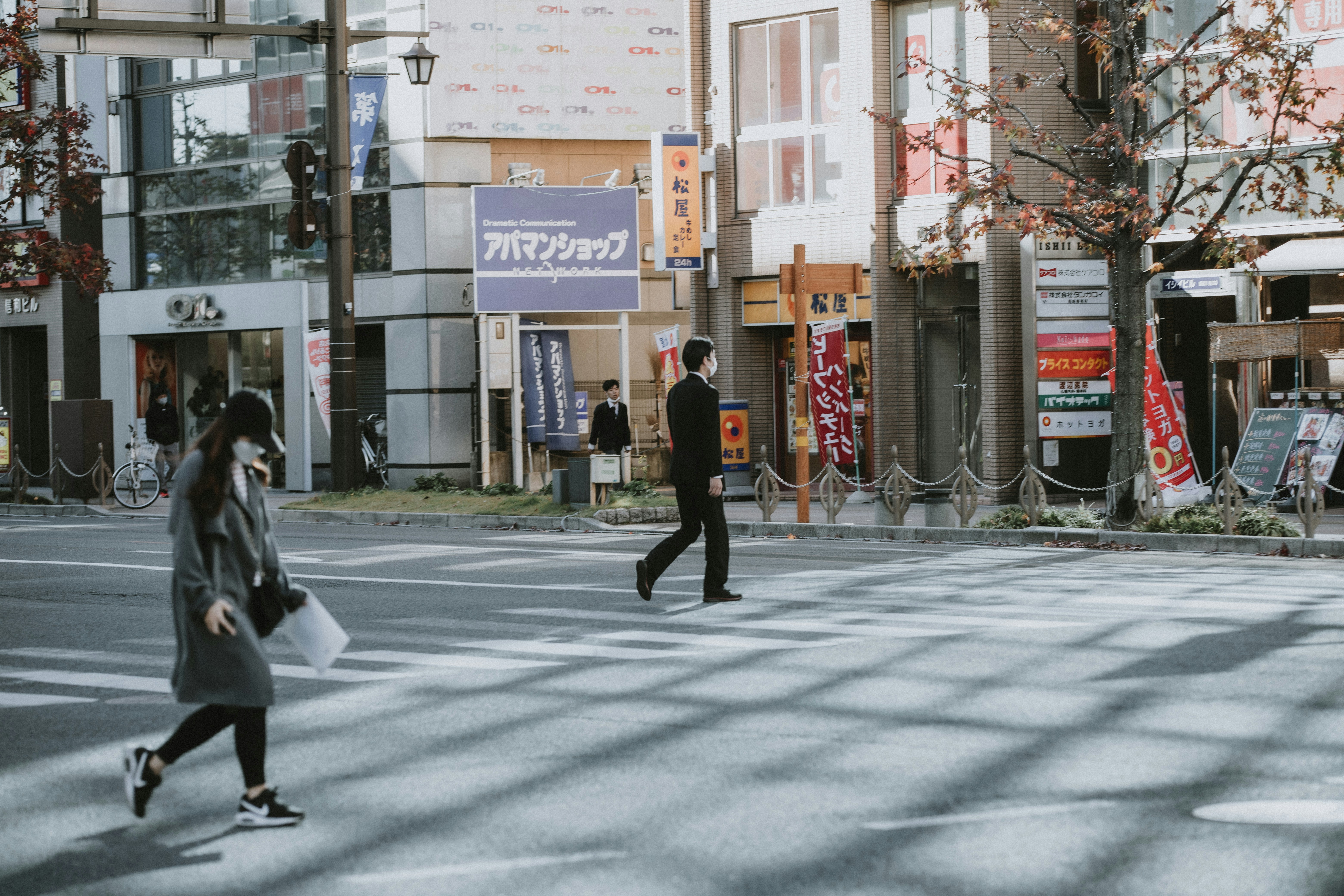 A man walks across a city street as a woman strides towards him, framed by shadows and storefronts. The scene captures the essence of urban life.