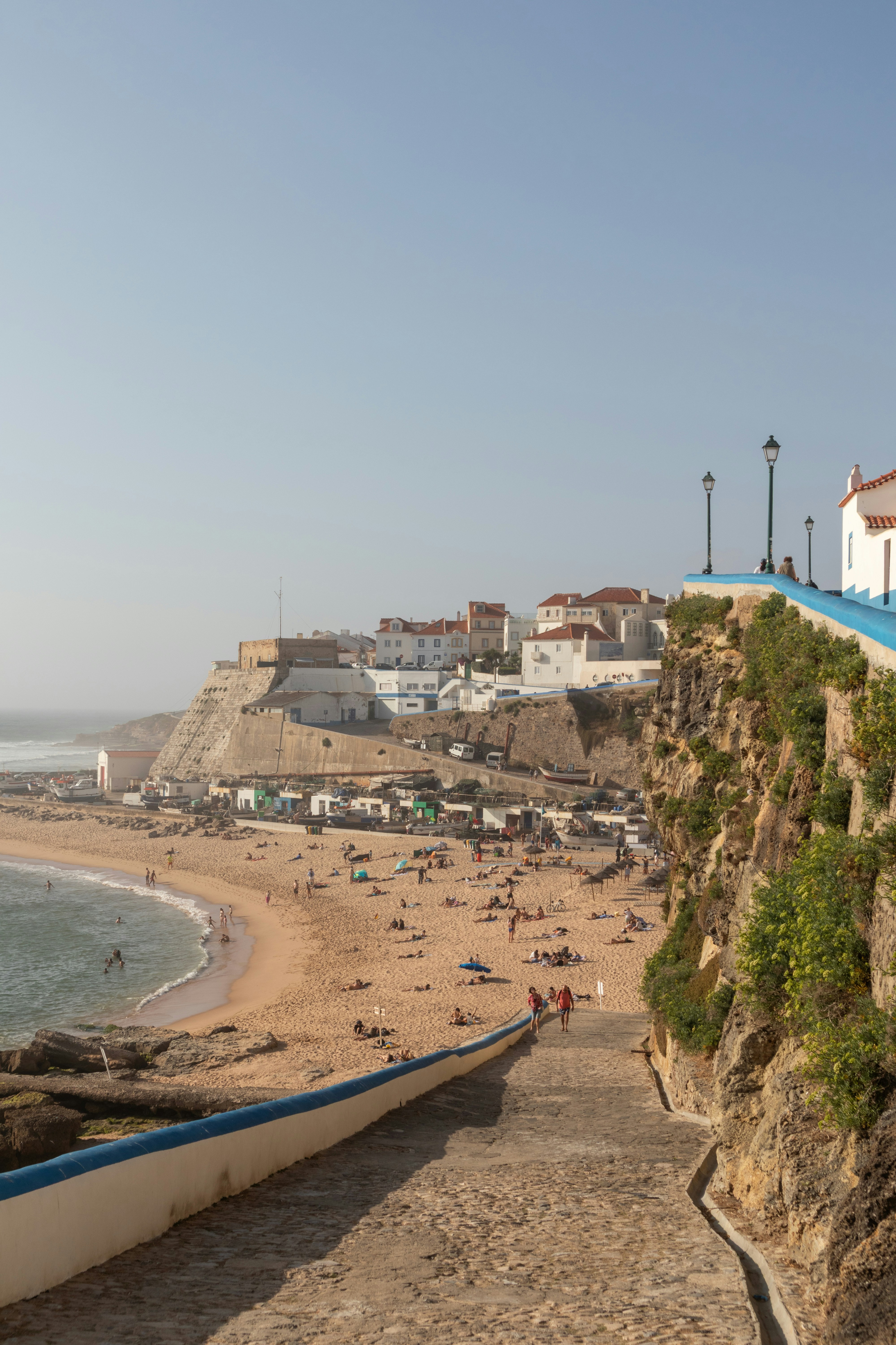 Winding path leading down to a sandy beach with sunbathers and coastal buildings in the background. The scene captures a tranquil beach atmosphere.