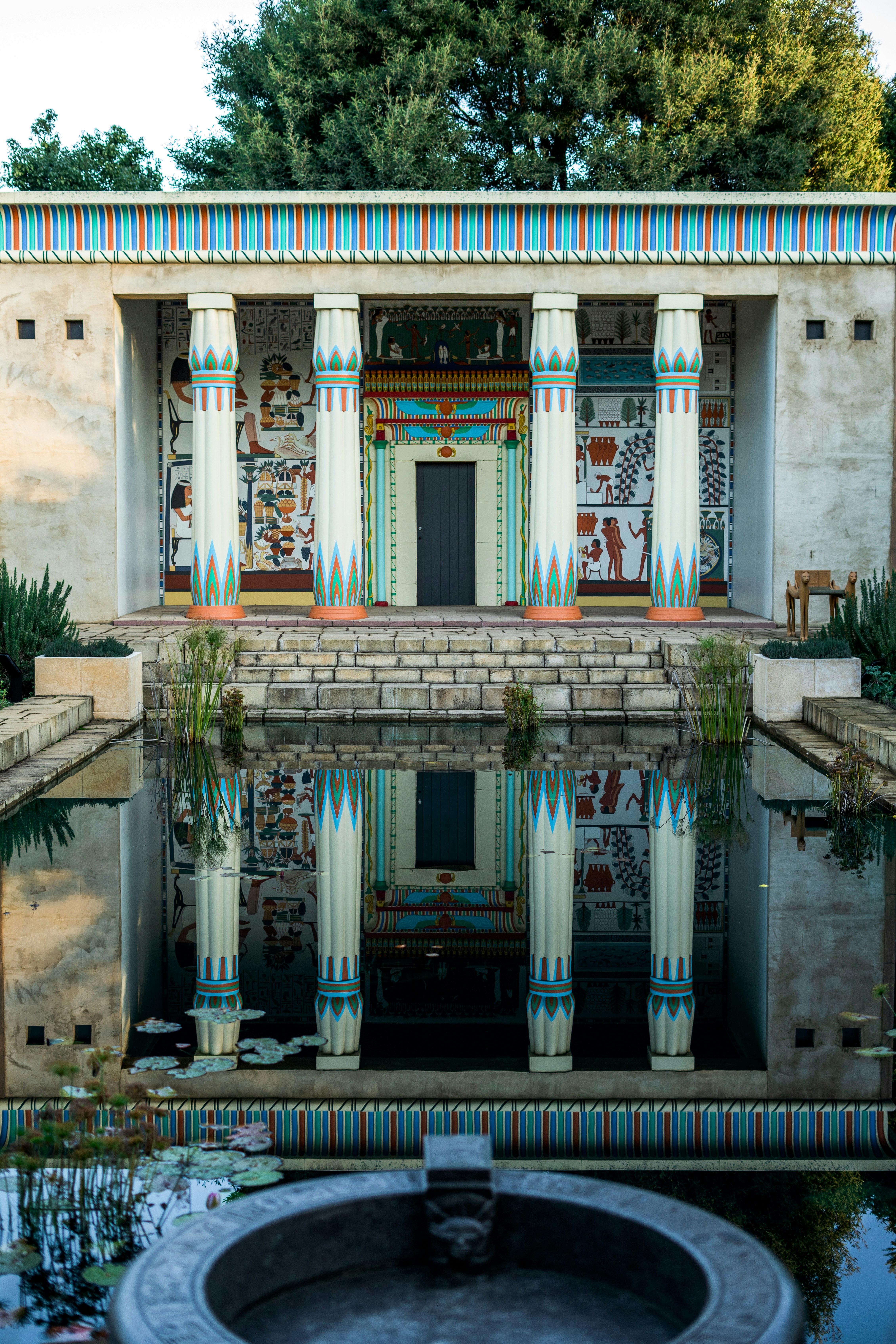 Vibrant temple facade adorned with intricate murals, reflected in a serene pond surrounded by lush greenery.