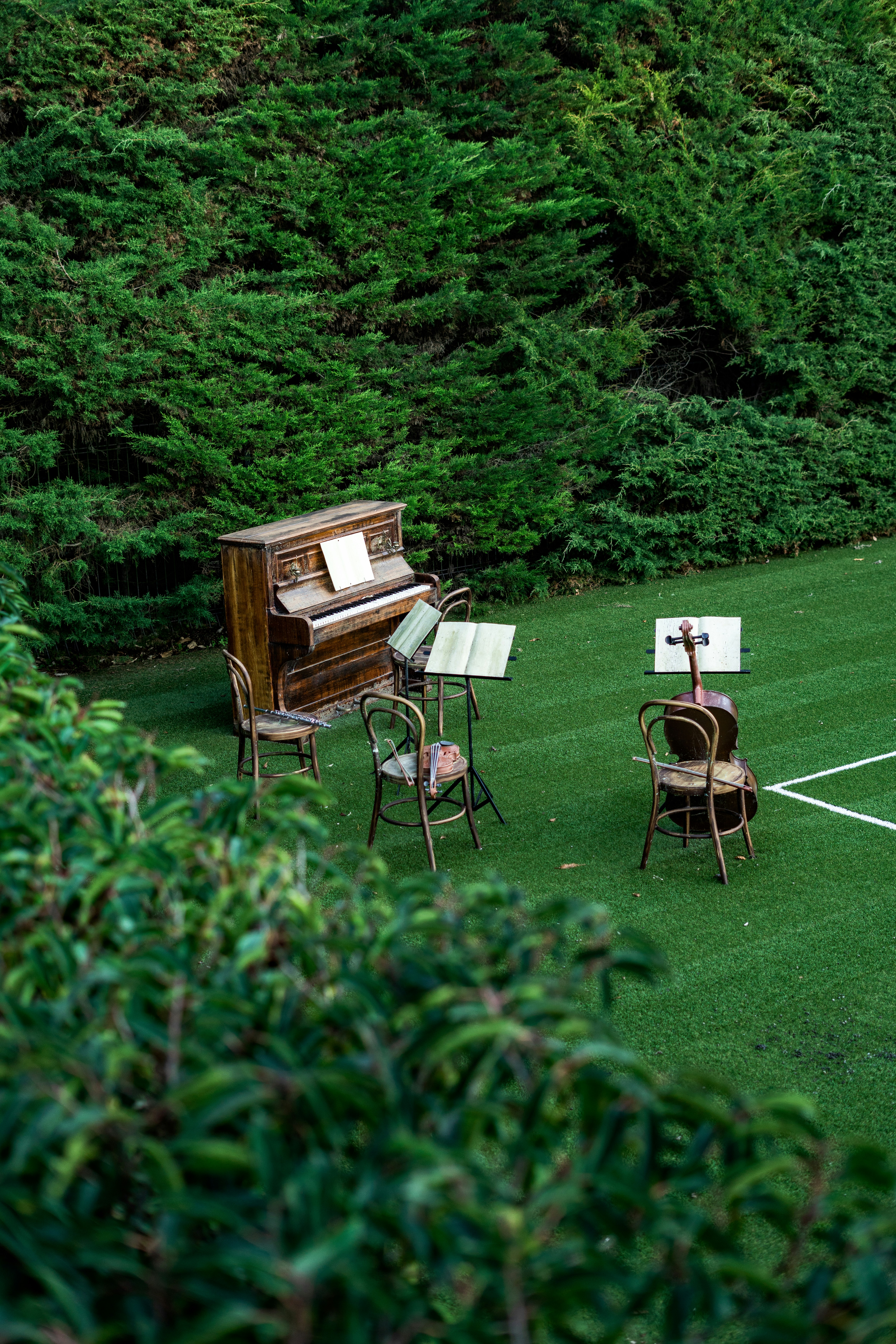 An outdoor music setup featuring an antique piano, chairs, and music stands surrounded by lush greenery. The scene invites a serene atmosphere for a musical gathering.